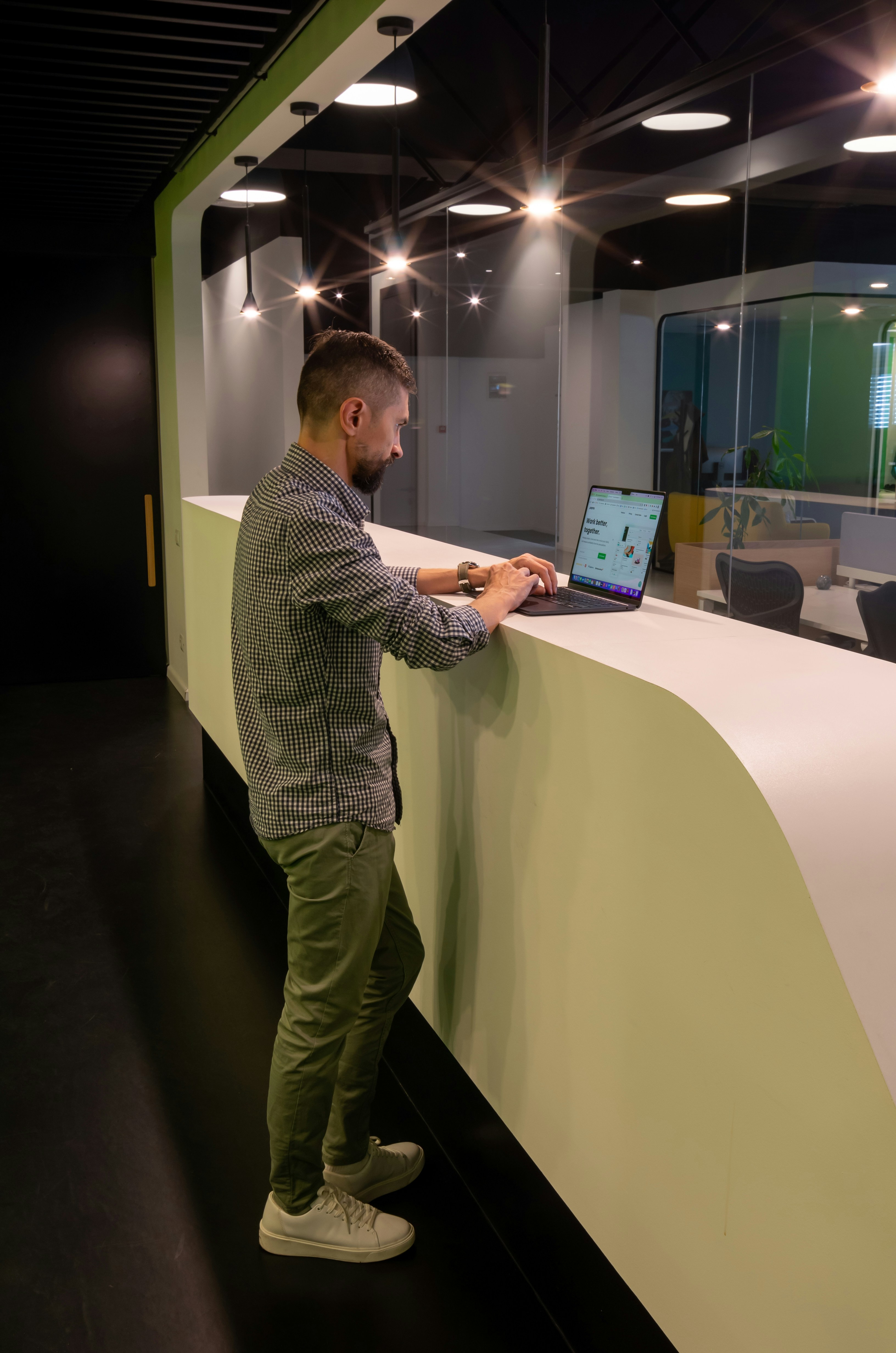 Man in a checkered shirt working intently on a laptop at a modern workspace, surrounded by glass partitions and contemporary design elements.