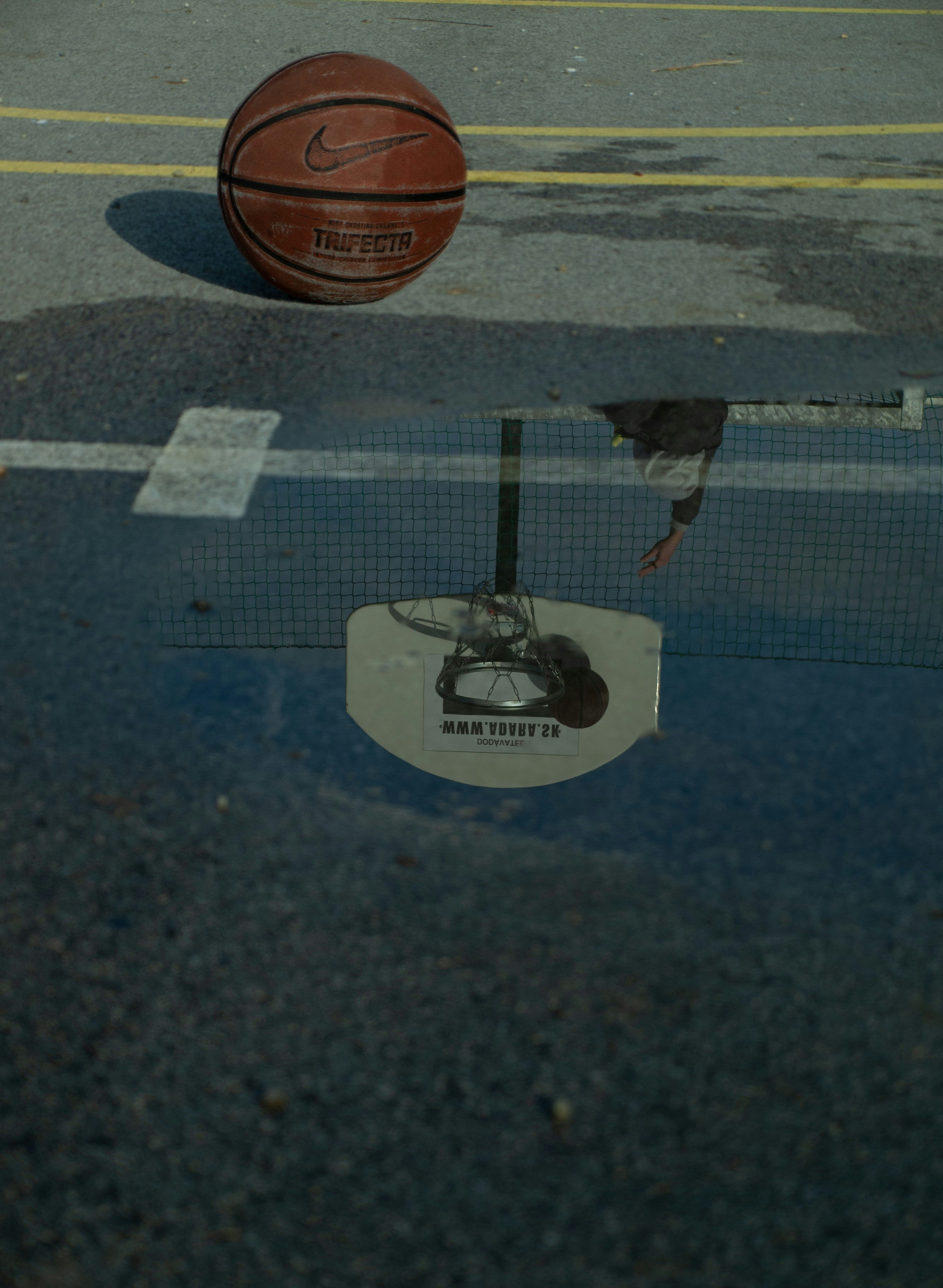 A basketball rests beside a puddle, reflecting a hoop and player in motion. The scene captures the essence of street basketball culture.