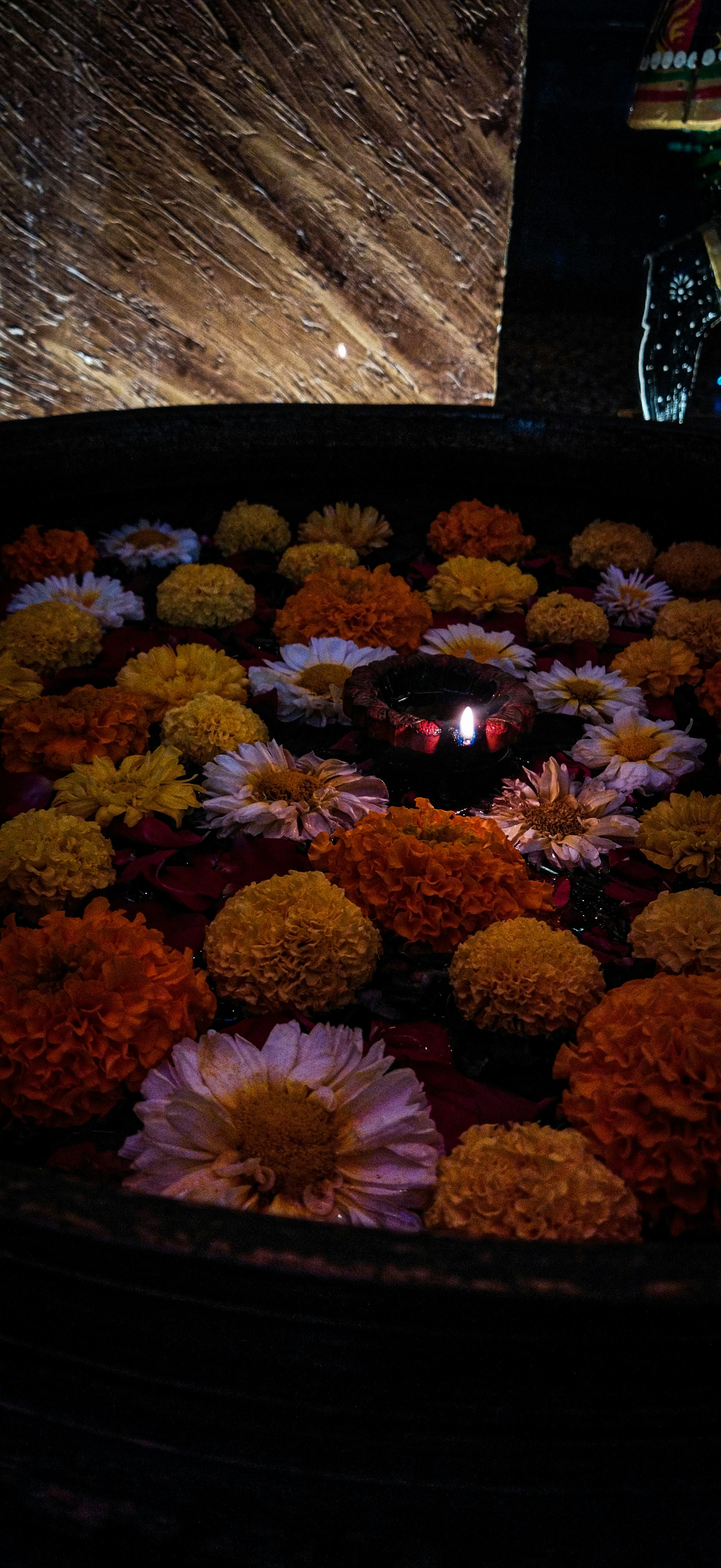 Floating marigold and daisy flowers surround a small lit diya in a darkened setting, creating a serene atmosphere.