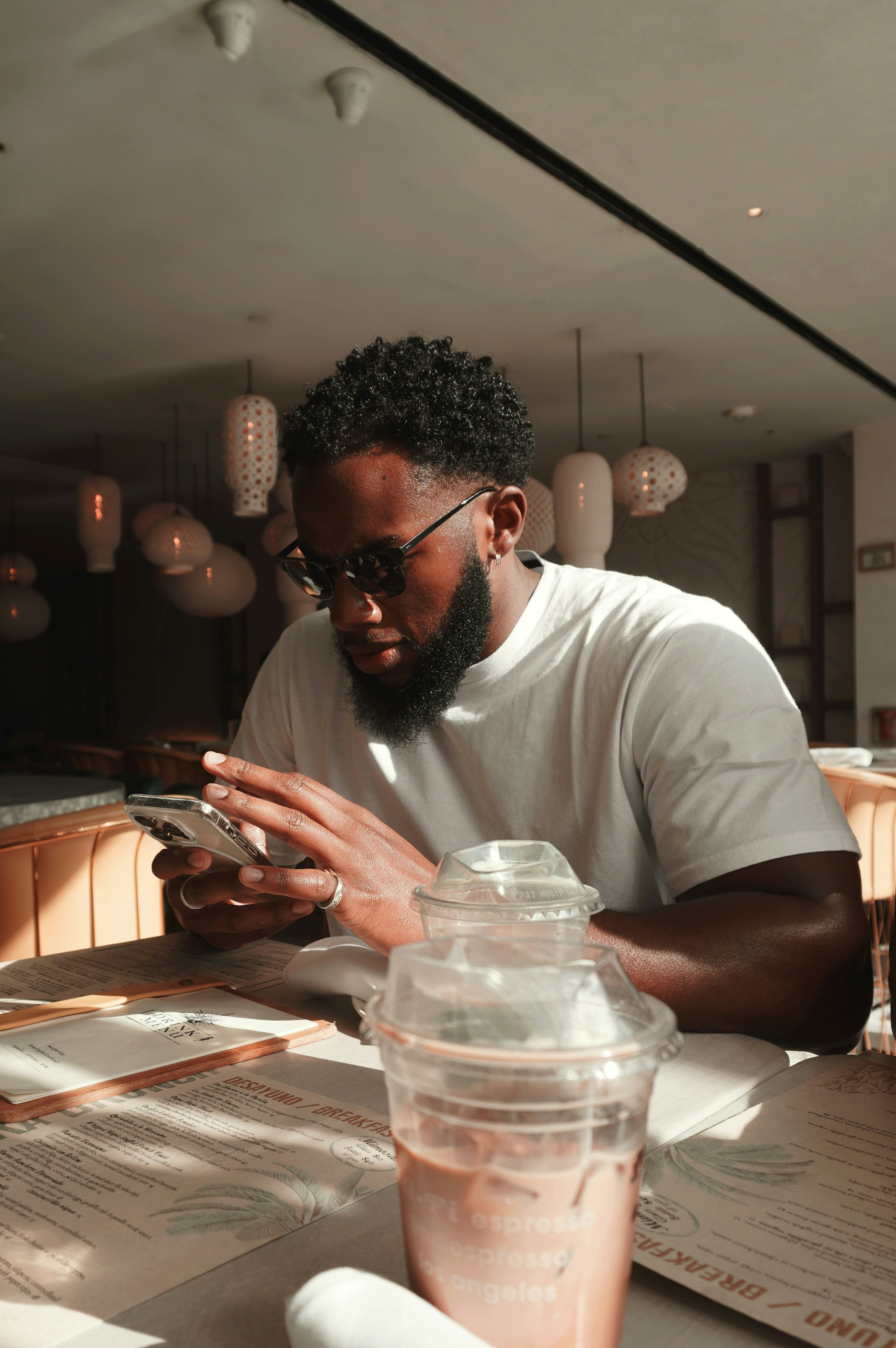 Bearded man in a white T-shirt sits at a sunlit cafe table, absorbed in his smartphone. A layered smoothie cup sits in the foreground.