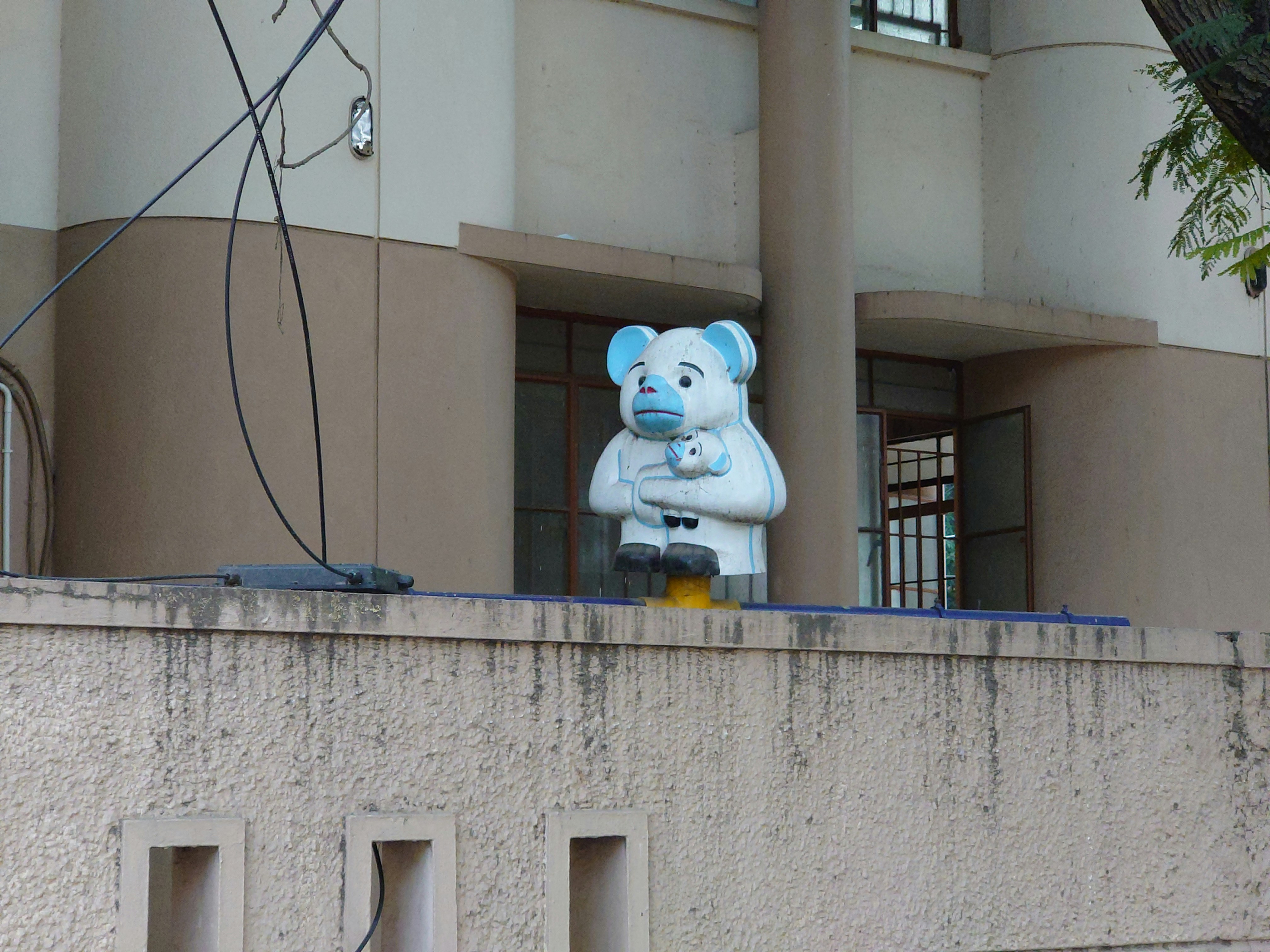 A charming bear sculpture holding a smaller bear, perched on a balcony of a building. The playful design adds character to the urban landscape.