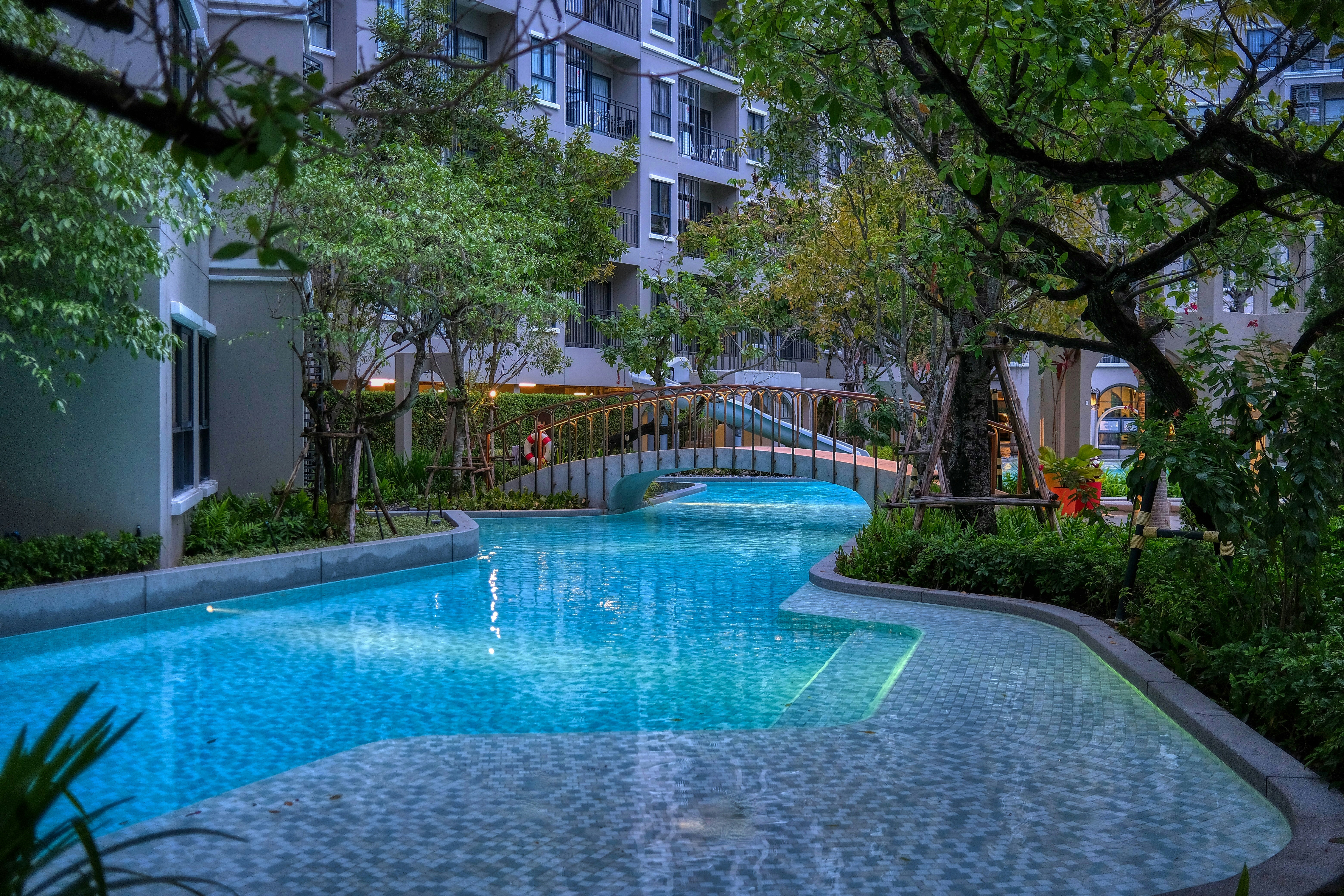 Curved swimming pool surrounded by lush greenery and modern apartment buildings under a soft evening glow.