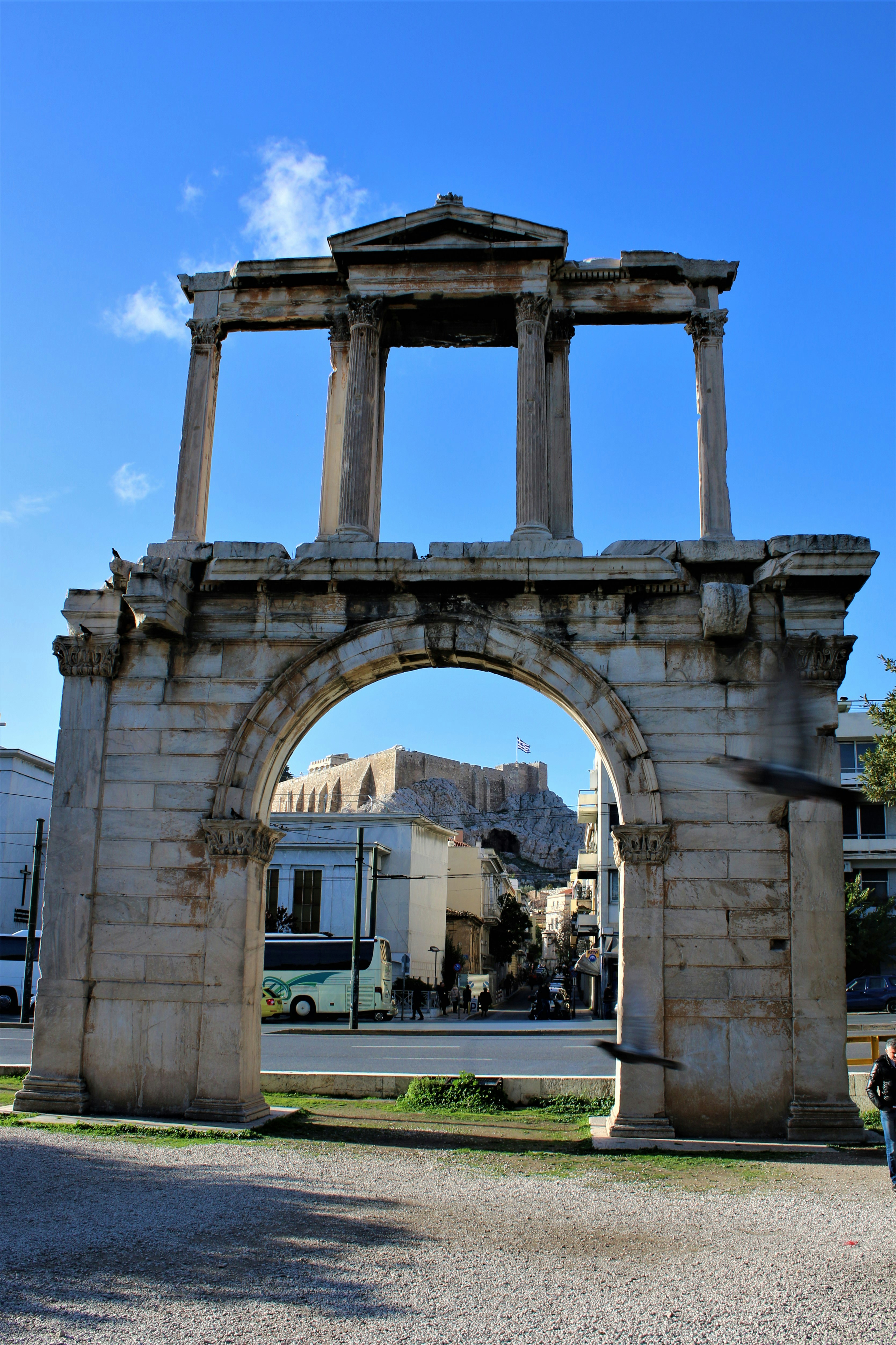 Ancient stone archway framing the Acropolis, with modern city life bustling beneath. The structure's intricate columns contrast with the contemporary surroundings.