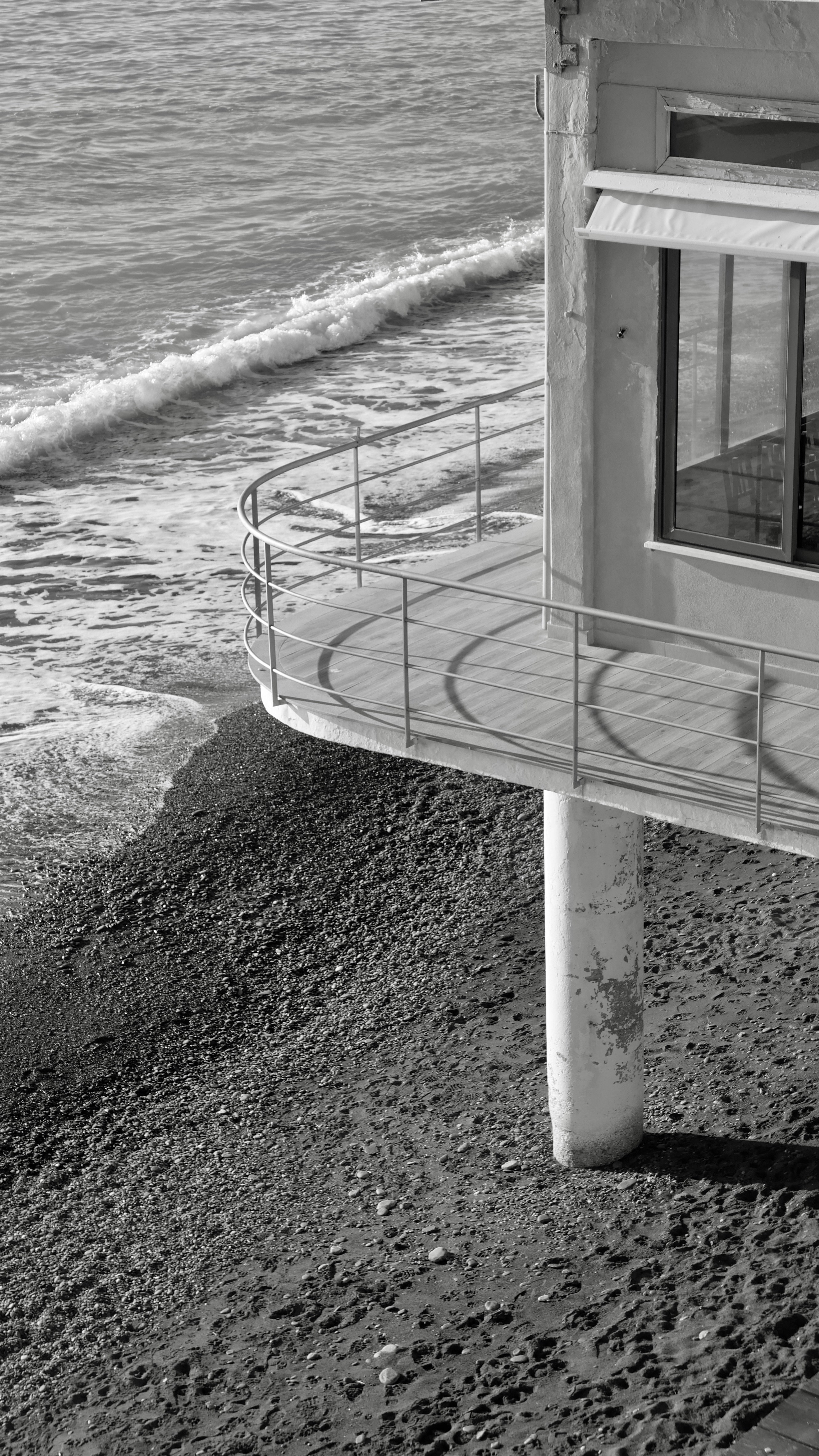 Monochrome photograph of a curved railing and coastal building perched above a pebbled beach, with waves breaking nearby.
