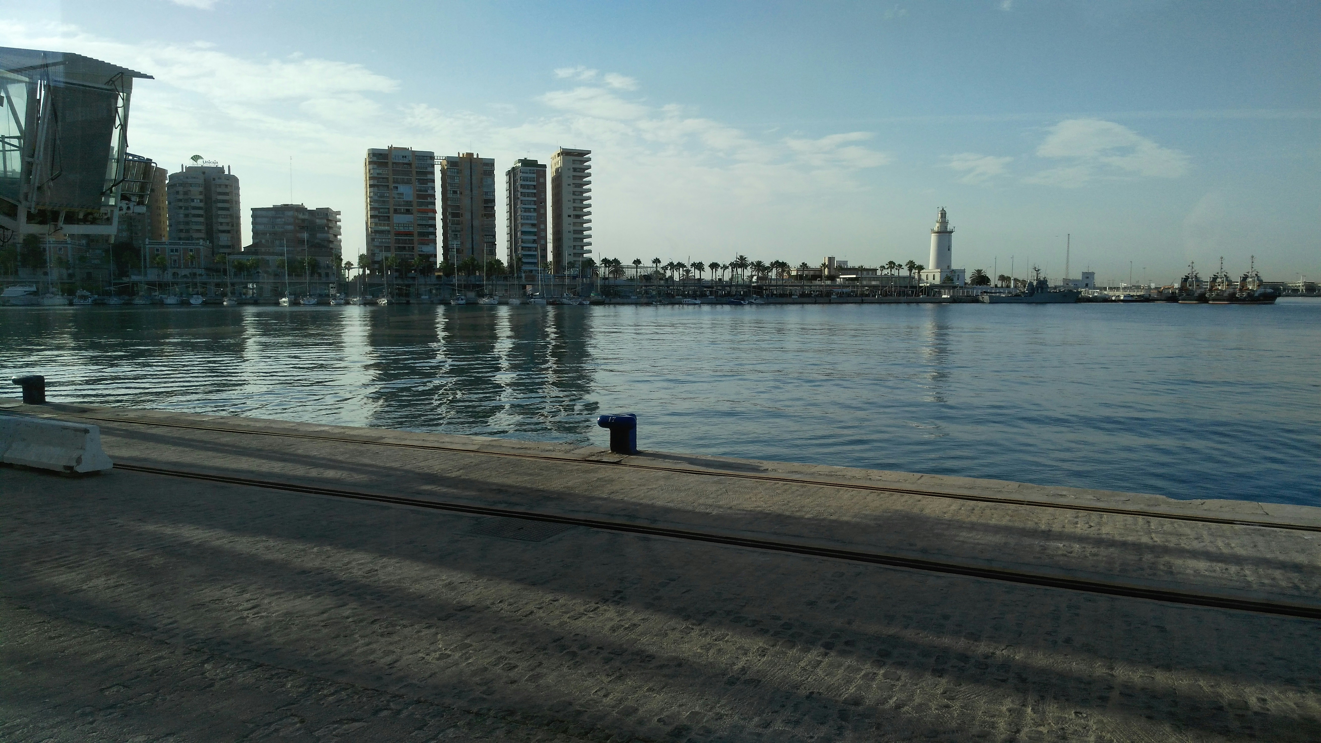Calm harbor scene with modern high-rises along the waterfront and a distant lighthouse across reflective water.