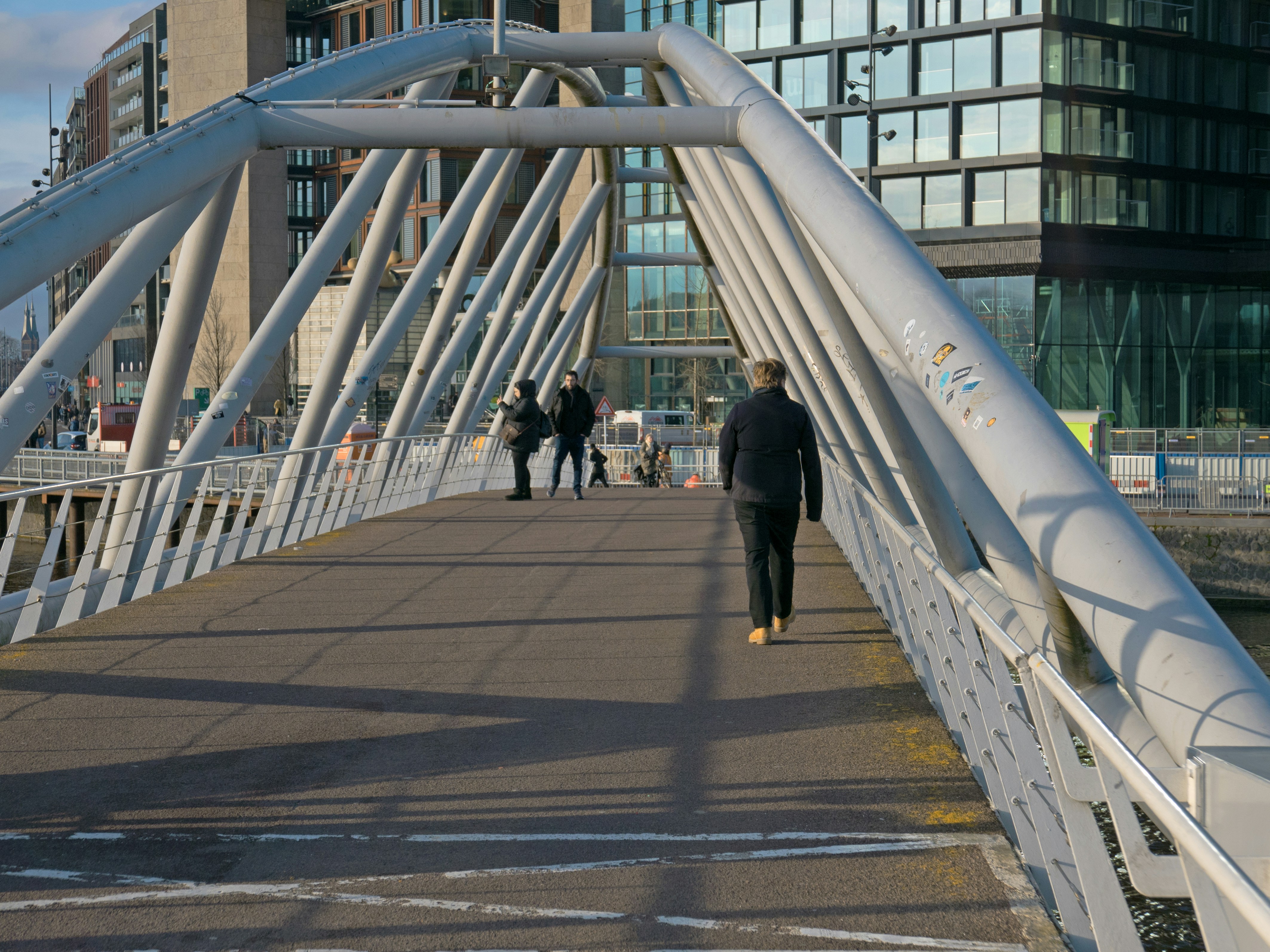 Photo of city people, walking over the metallic foot bridge over the Oosterdok water in Amsterdam city - a modern steel construction & glass architecture building facades in the background. It is a sunny day in winter. Free photo of human street photography in Amsterdam, Netherlands by Fons Heijnsbroek. / Foto van mensen, lopend in de moderne staalconstructie van de J.J. van der Veldebrug, over het Oosterdok - hij verbindt het Nemo Science Museum met het Oosterdokseiland - moderne architectuur te Amsterdam - straatfotografie Nederland in hoge resolutie foto, Fons Heijnsbroek; gratis download,