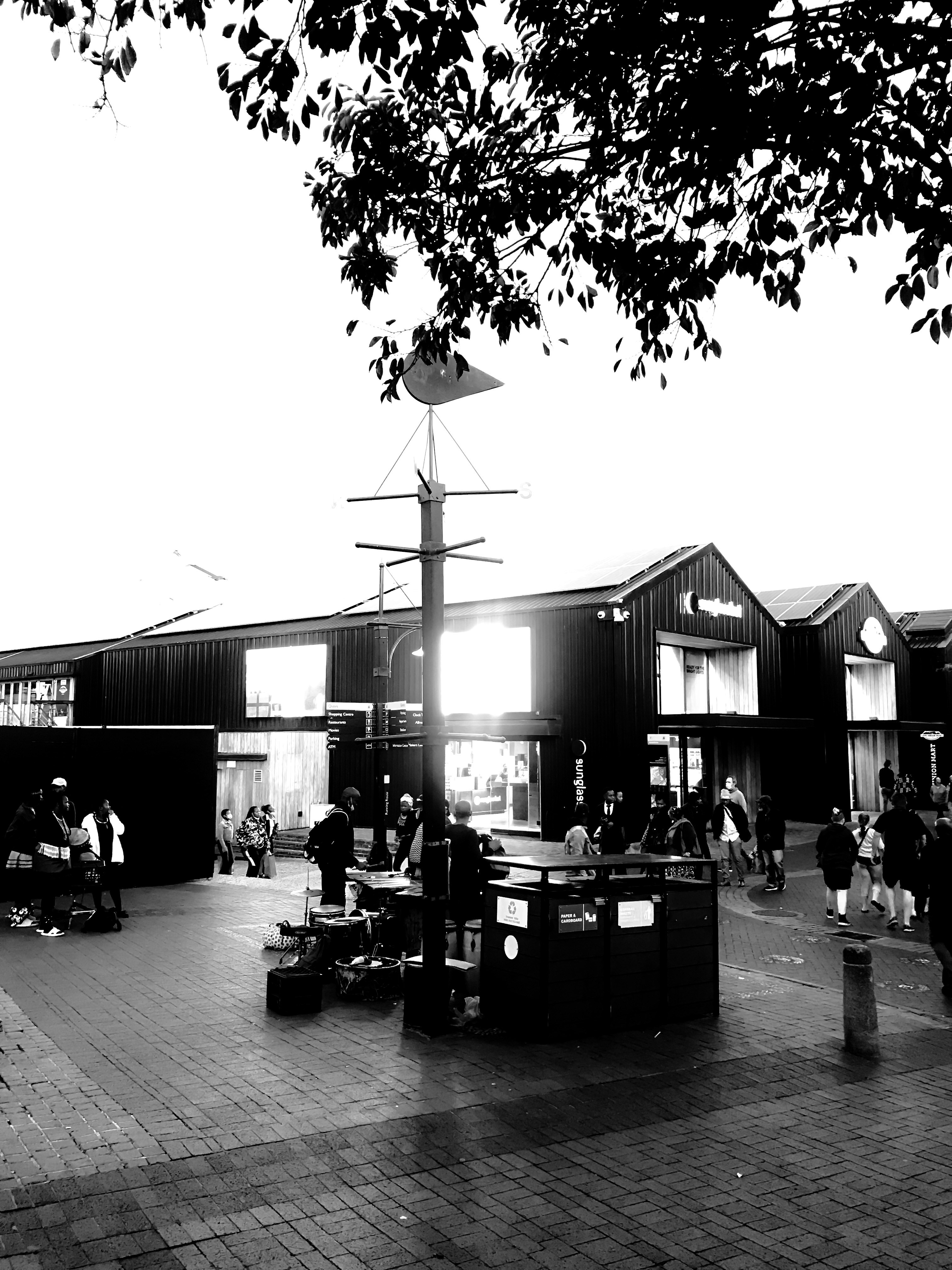 A lively urban scene featuring people congregating near food stalls and shops, framed by modern architecture and trees. The monochrome effect adds a timeless quality.