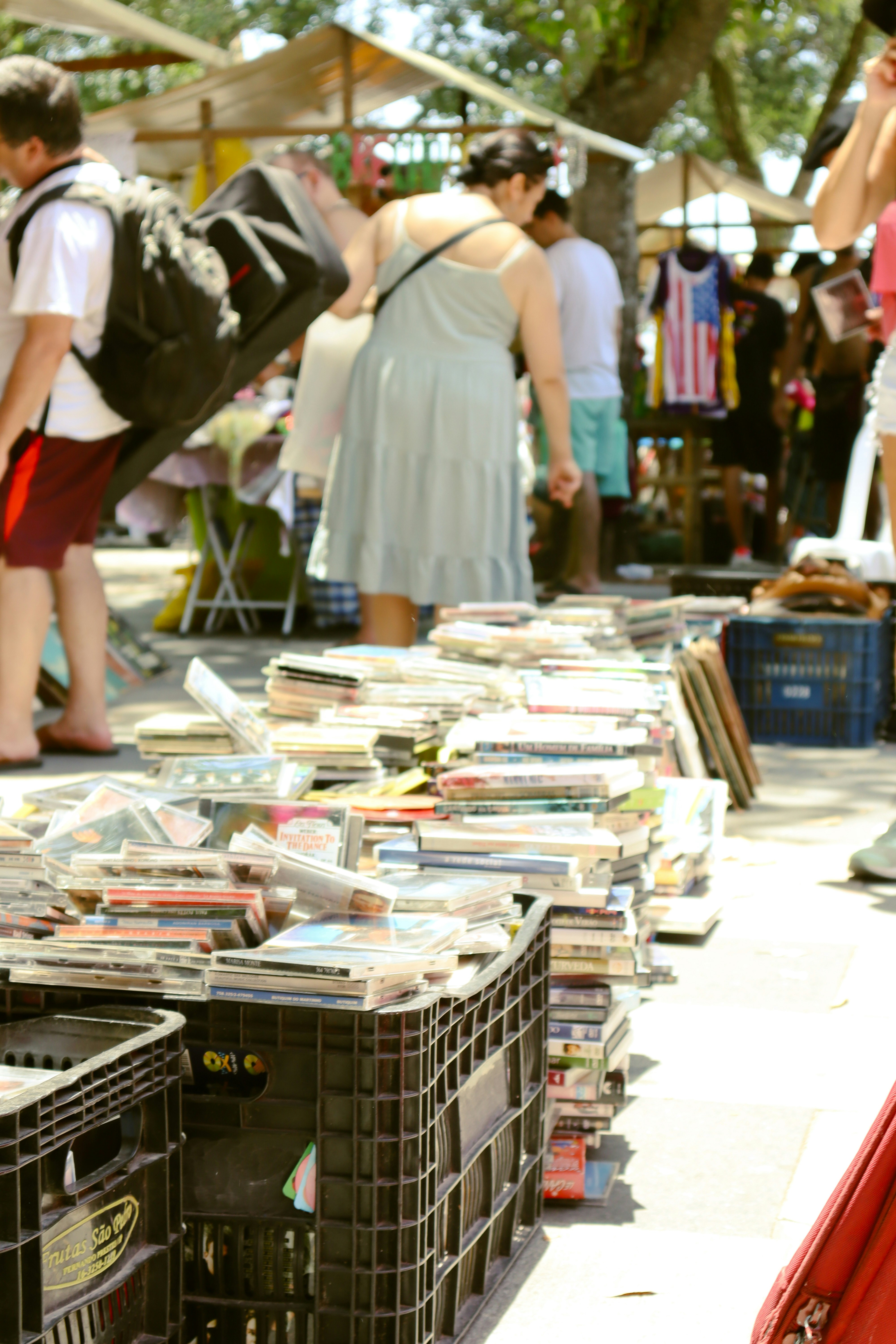 Outdoor market scene with stacks of books arranged on crates in the foreground and shoppers under canopies in the background.