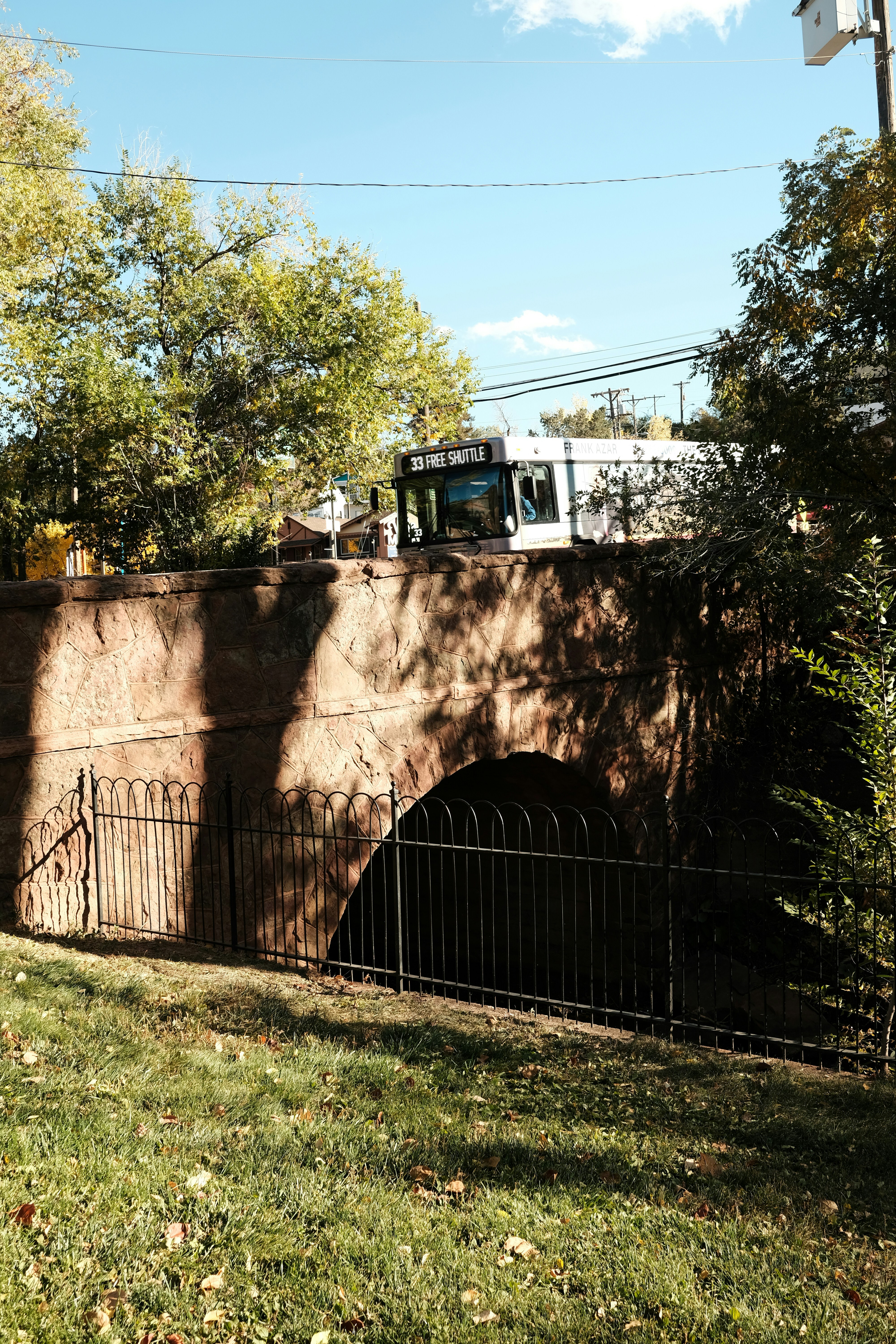 A bus labeled '33 FREE SHUTTLE' parked near a stone bridge, surrounded by autumn foliage and urban infrastructure.