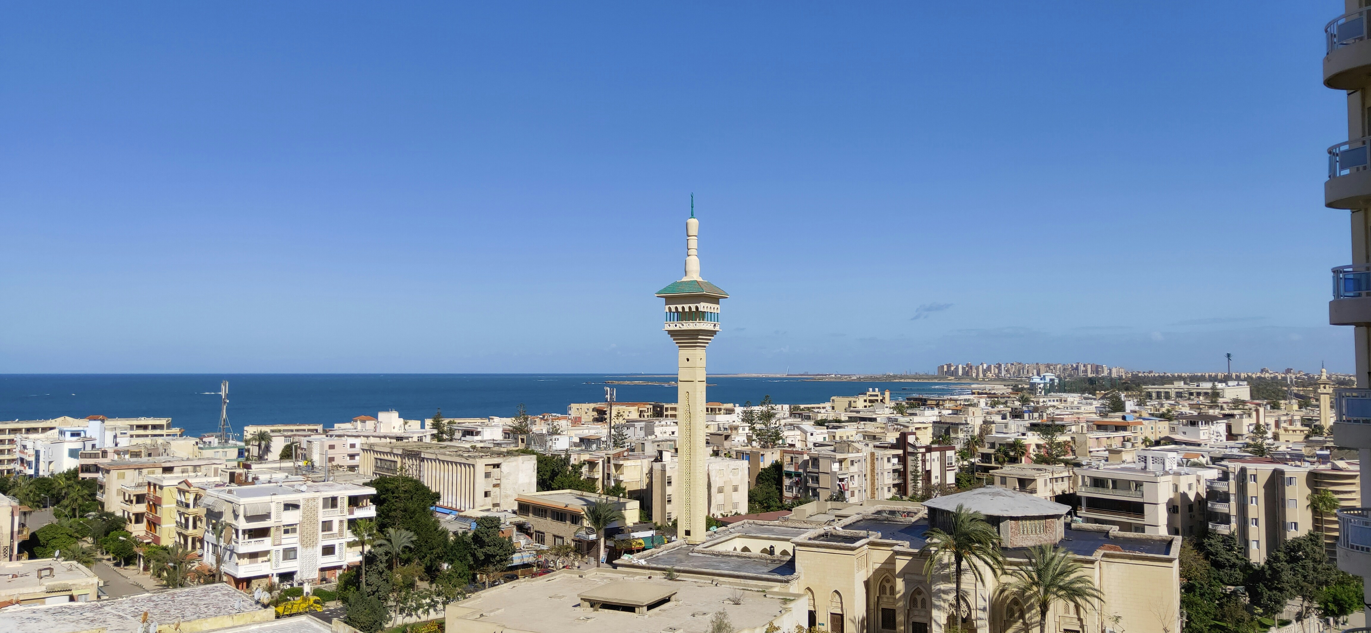 Minaret rising above a coastal city with clear blue skies and distant sea.