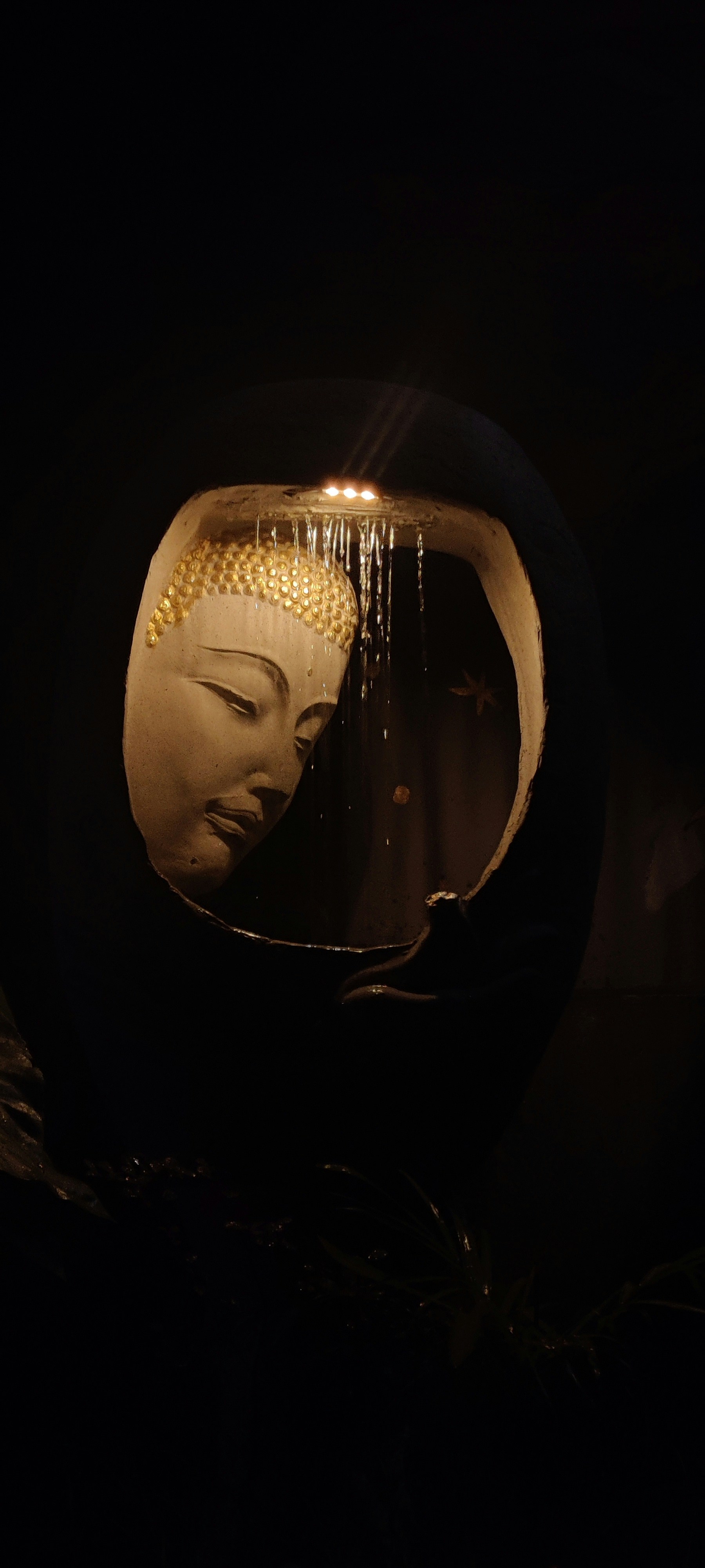 Dimly lit statue of a Buddha head behind a curved mask, with warm tones and a subtle waterfall glow; a contemplative shrine scene. Logo/pedestal not visible.