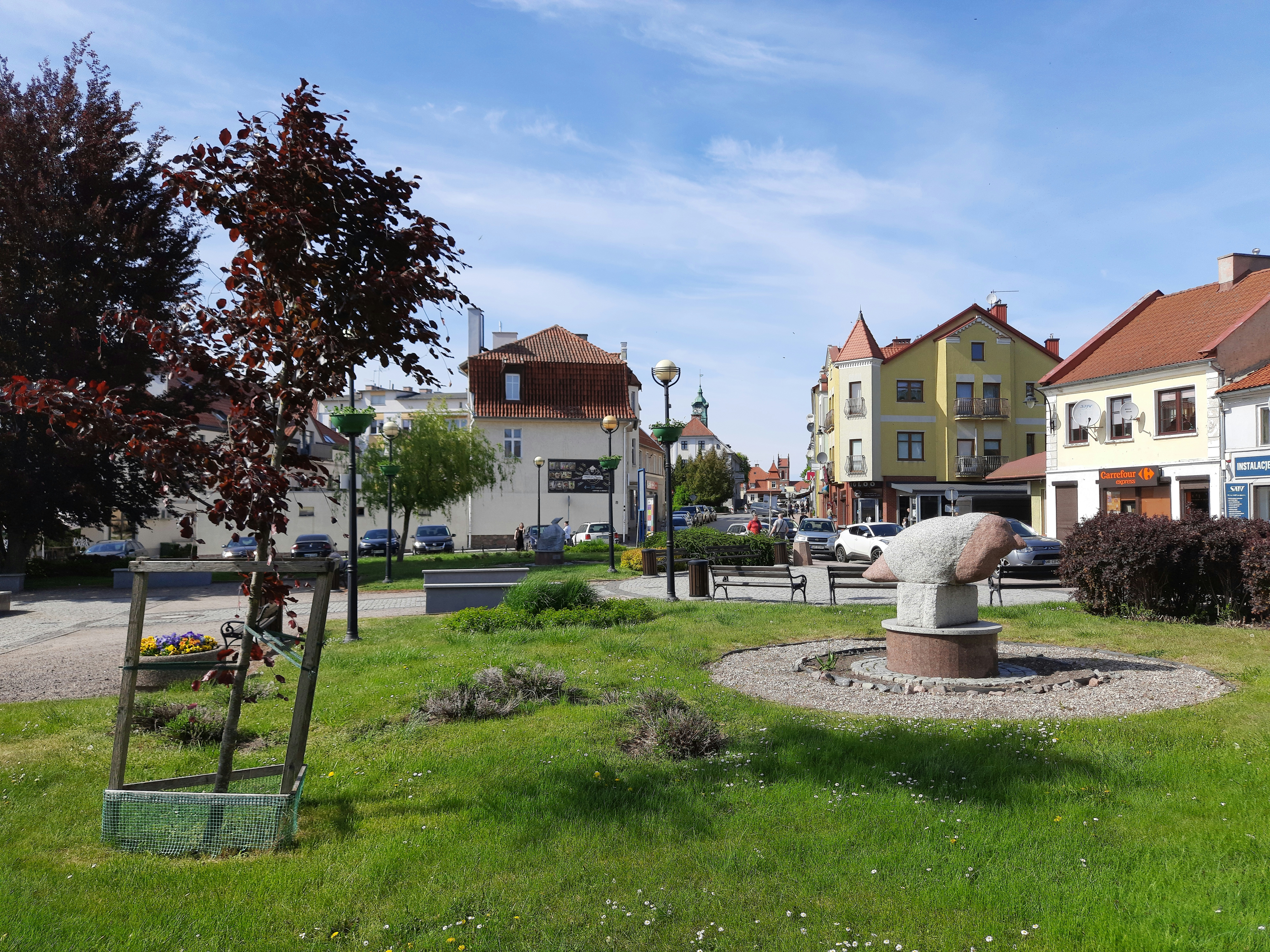 Sunlit town park scene with a circular garden, a central stone sculpture, benches, and pastel houses under a blue sky.