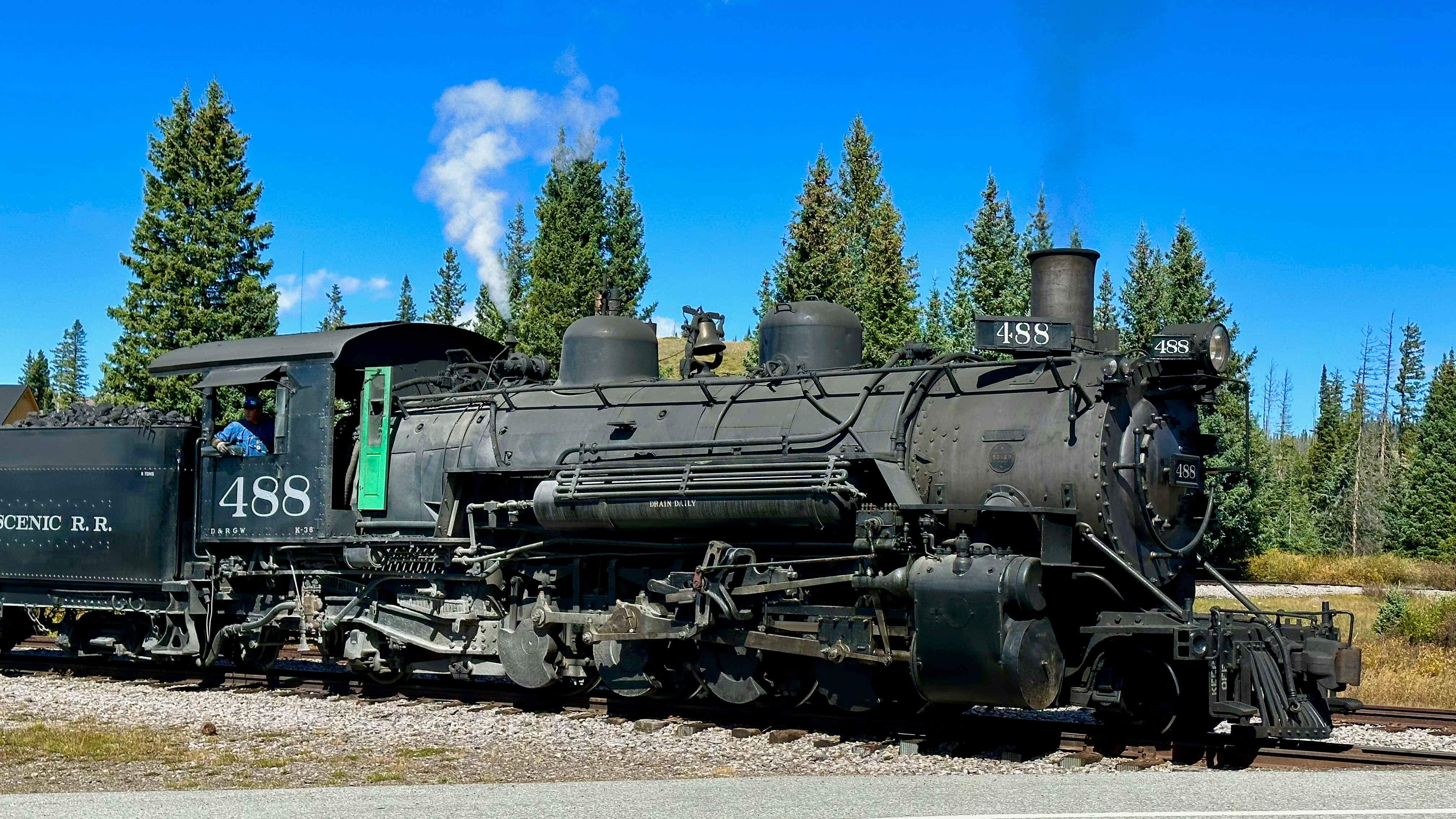 Historic steam locomotive 488 chugs along the scenic route, surrounded by lush evergreen trees and a clear blue sky.
