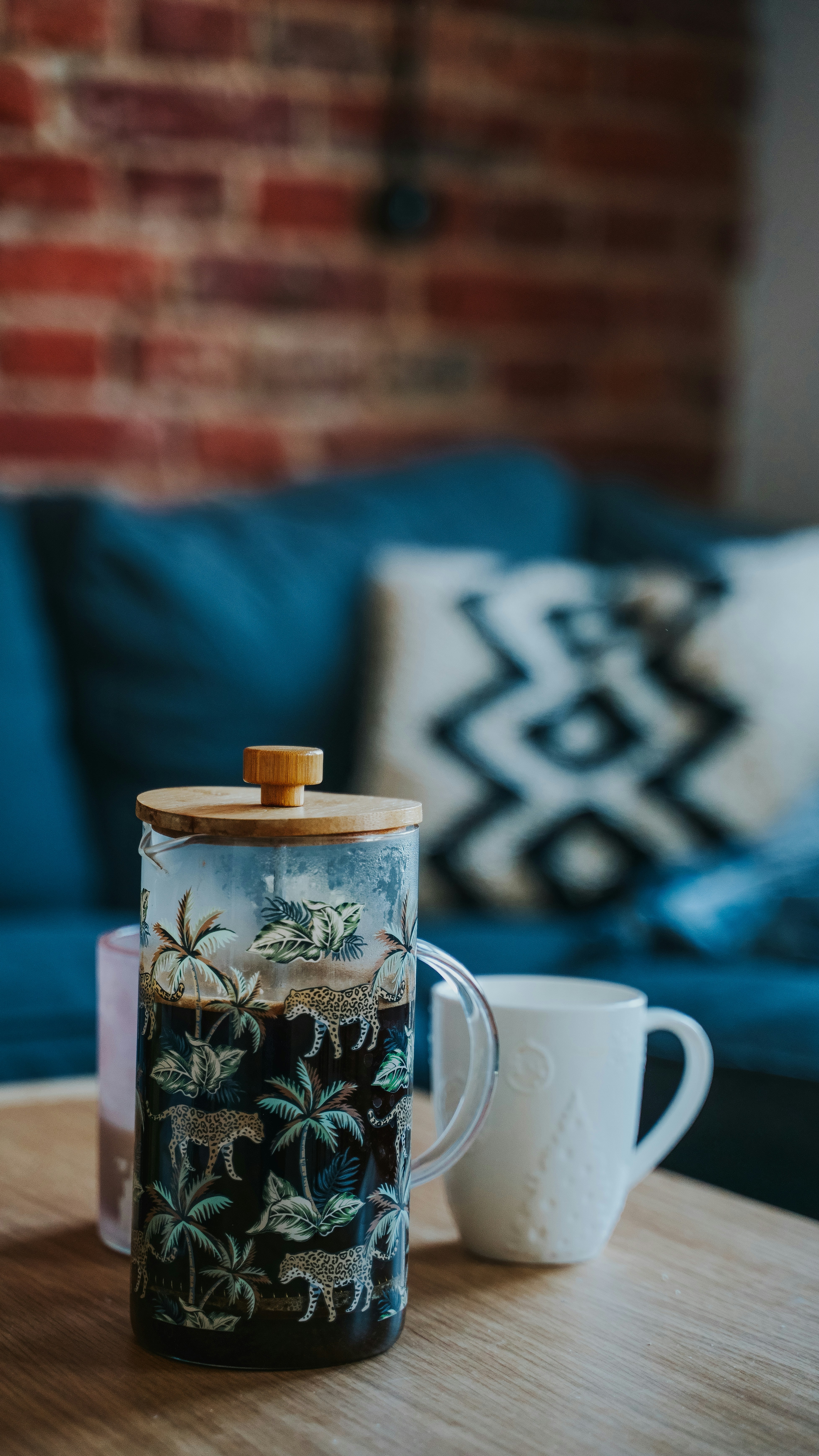 French press adorned with tropical motifs sits on a wooden table, accompanied by a white mug and a pastel glass. Cozy living room backdrop enhances the inviting atmosphere.