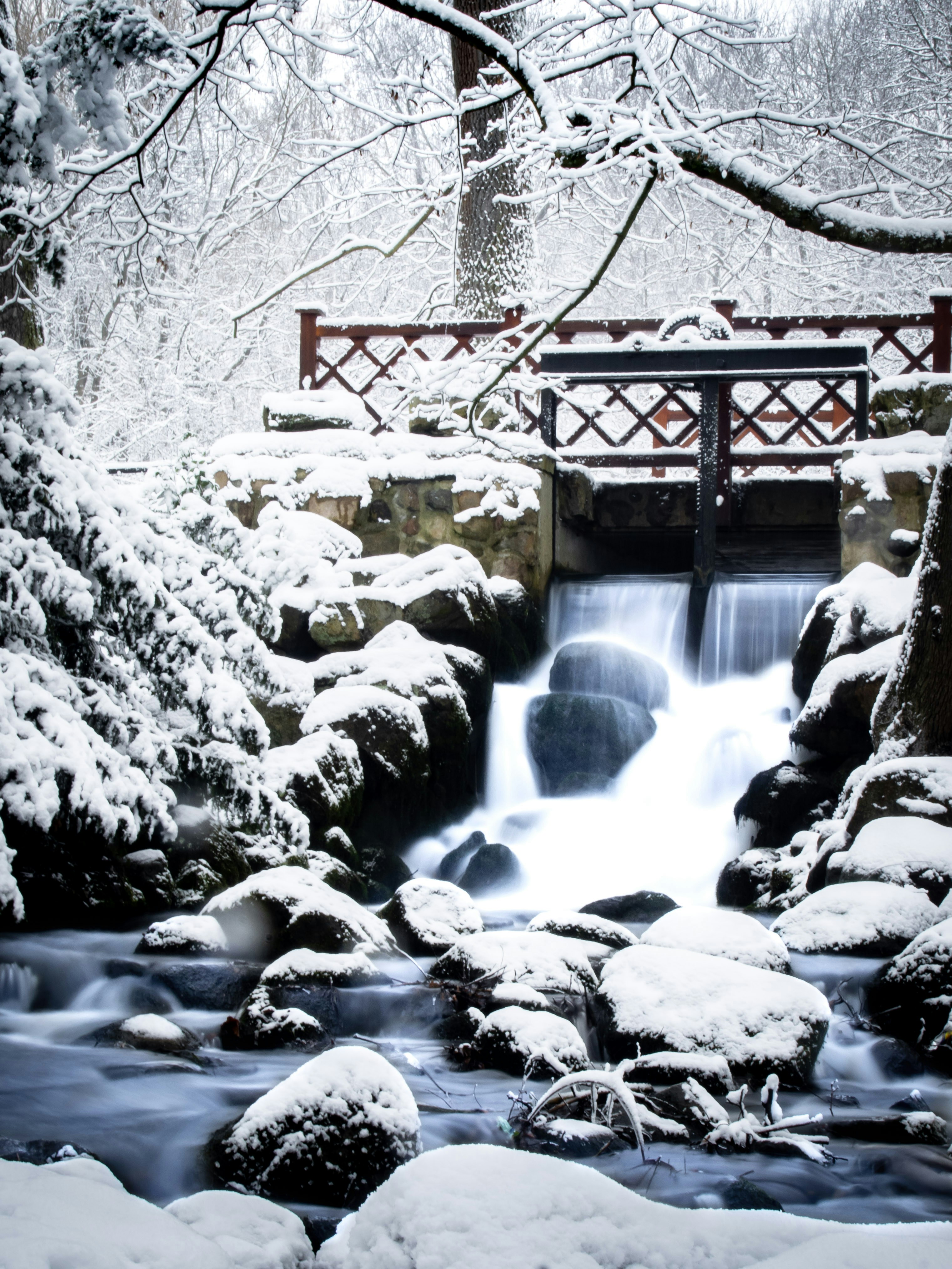 Snow-covered stones and a gentle stream flow beneath a rustic bridge in a serene winter landscape.