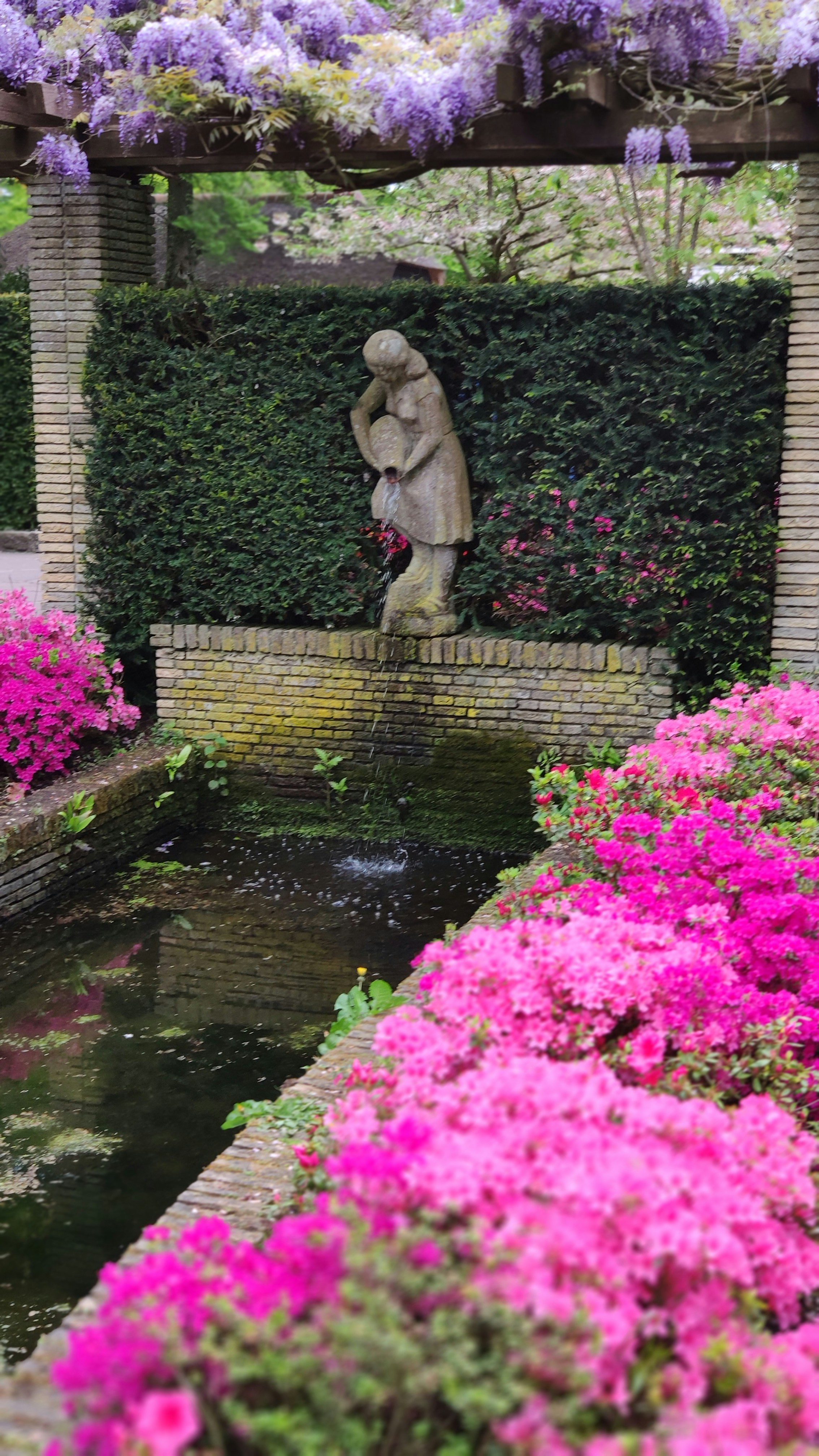 Stone statue of a mother cradling a child stands at a brick water feature, framed by a trimmed hedge and cascading purple wisteria with bright pink azaleas in the foreground.