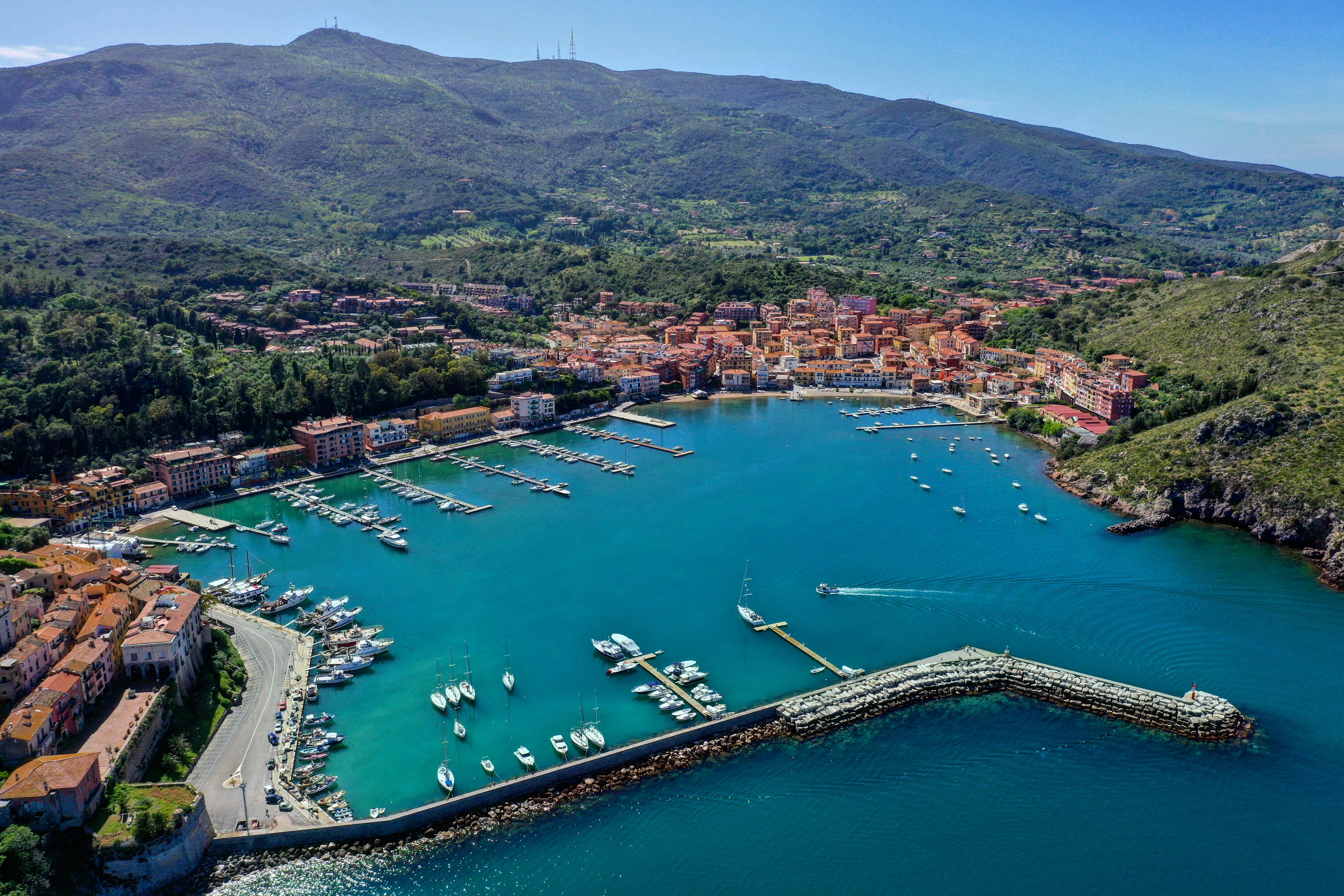 Aerial view of a vibrant harbor with boats docked along a winding shoreline, surrounded by lush green hills under a clear blue sky.