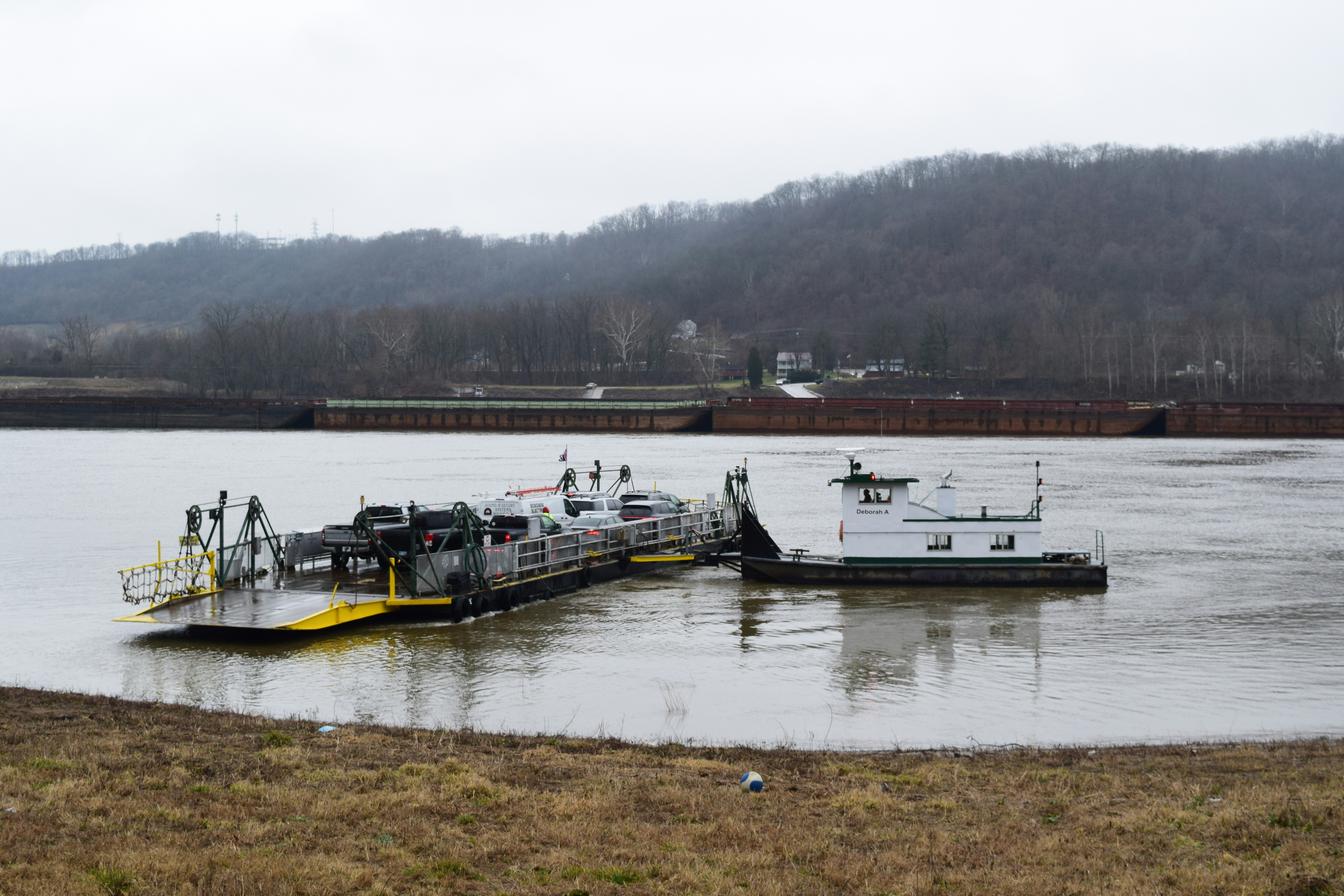 Small ferry loaded with vehicles traverses a calm river under overcast skies.
