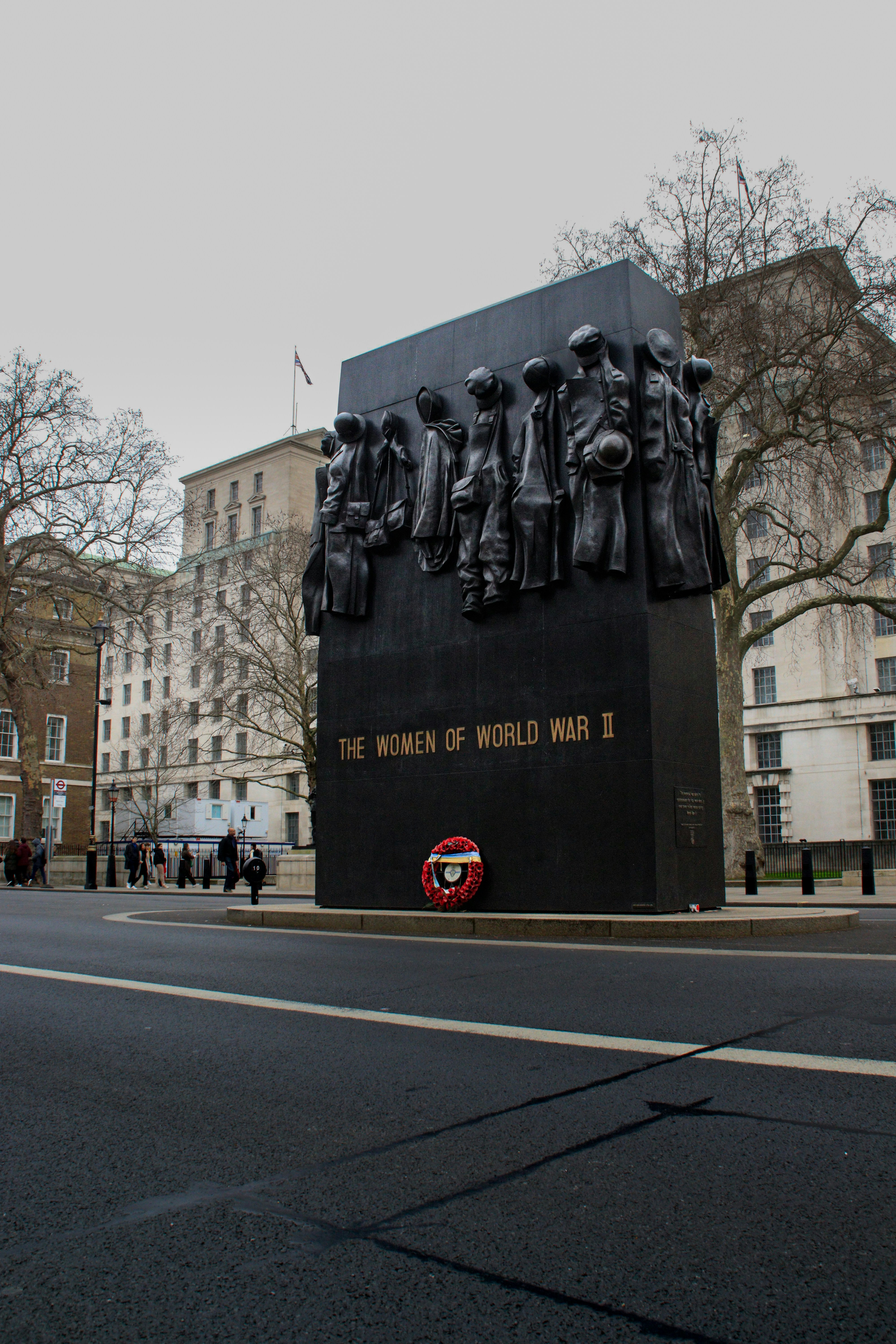 A large black memorial sculpture of women from World War II stands on a city street, with a red wreath laid at its base.