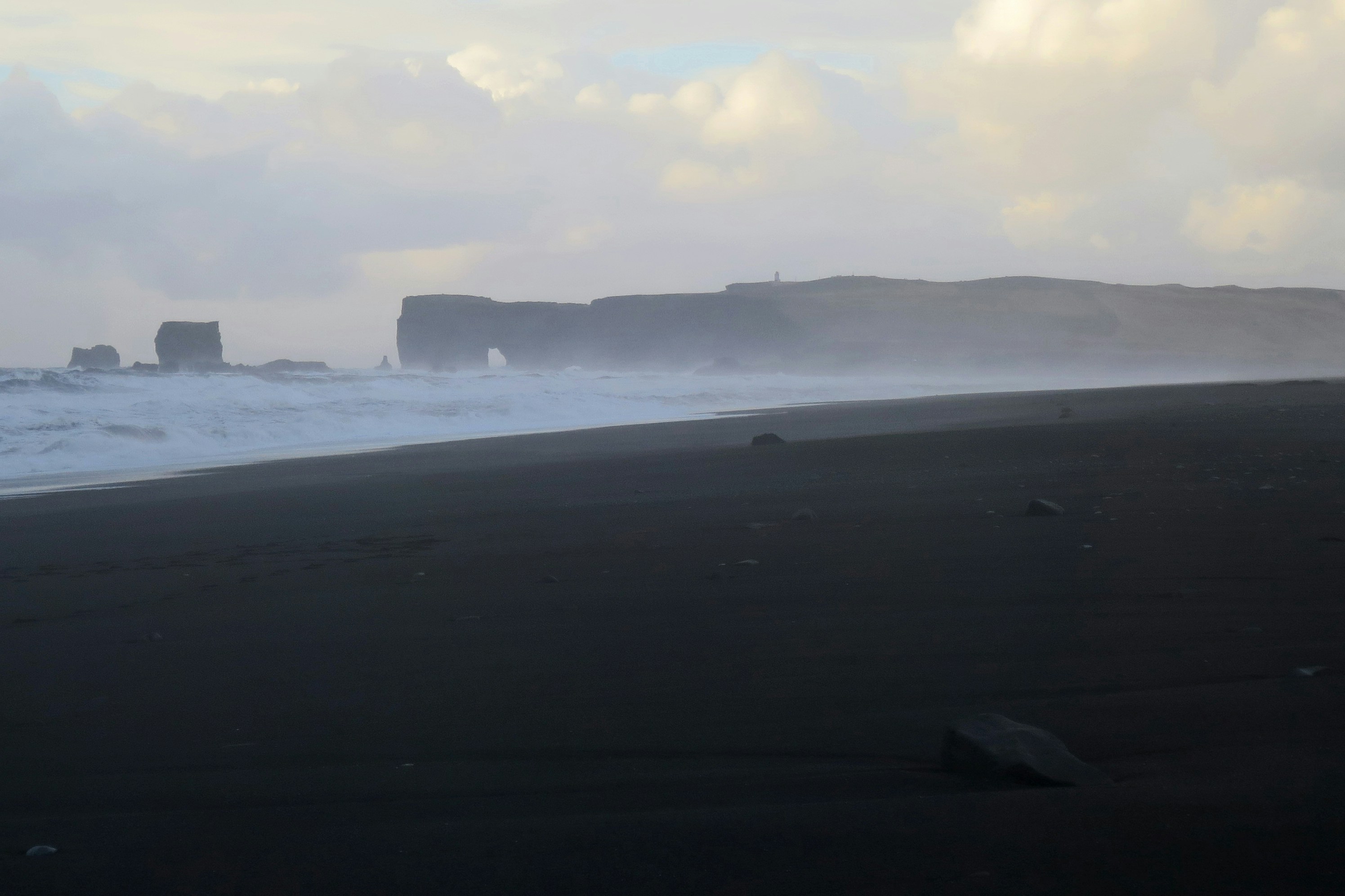 Waves crash against a dark sandy beach, with dramatic cliffs silhouetted against a cloudy sky. The scene evokes a sense of solitude and the raw power of nature.