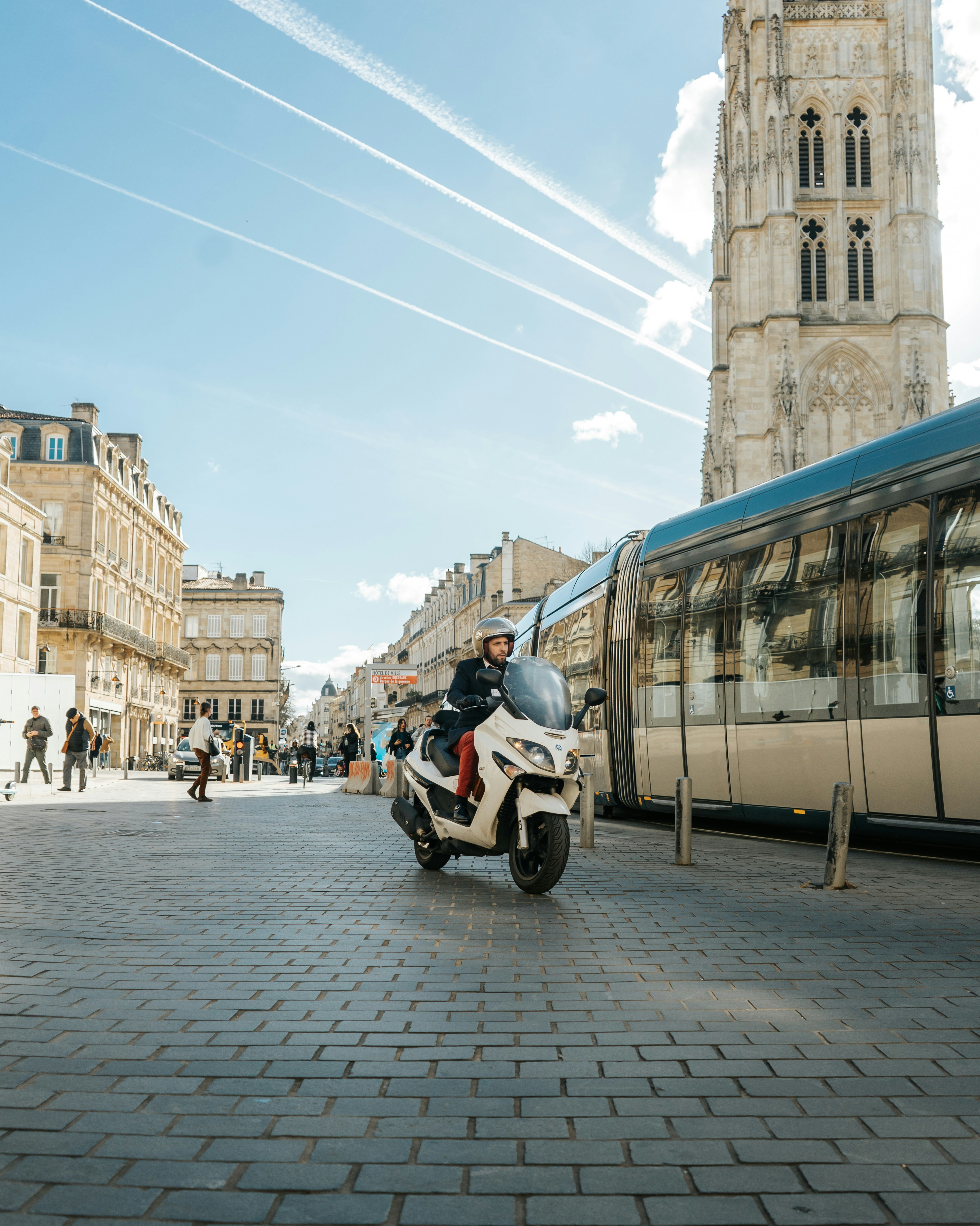 Rider on a White Scooter Navigates Sunlit Cobblestones beside a Modern Tram and Gothic Spire