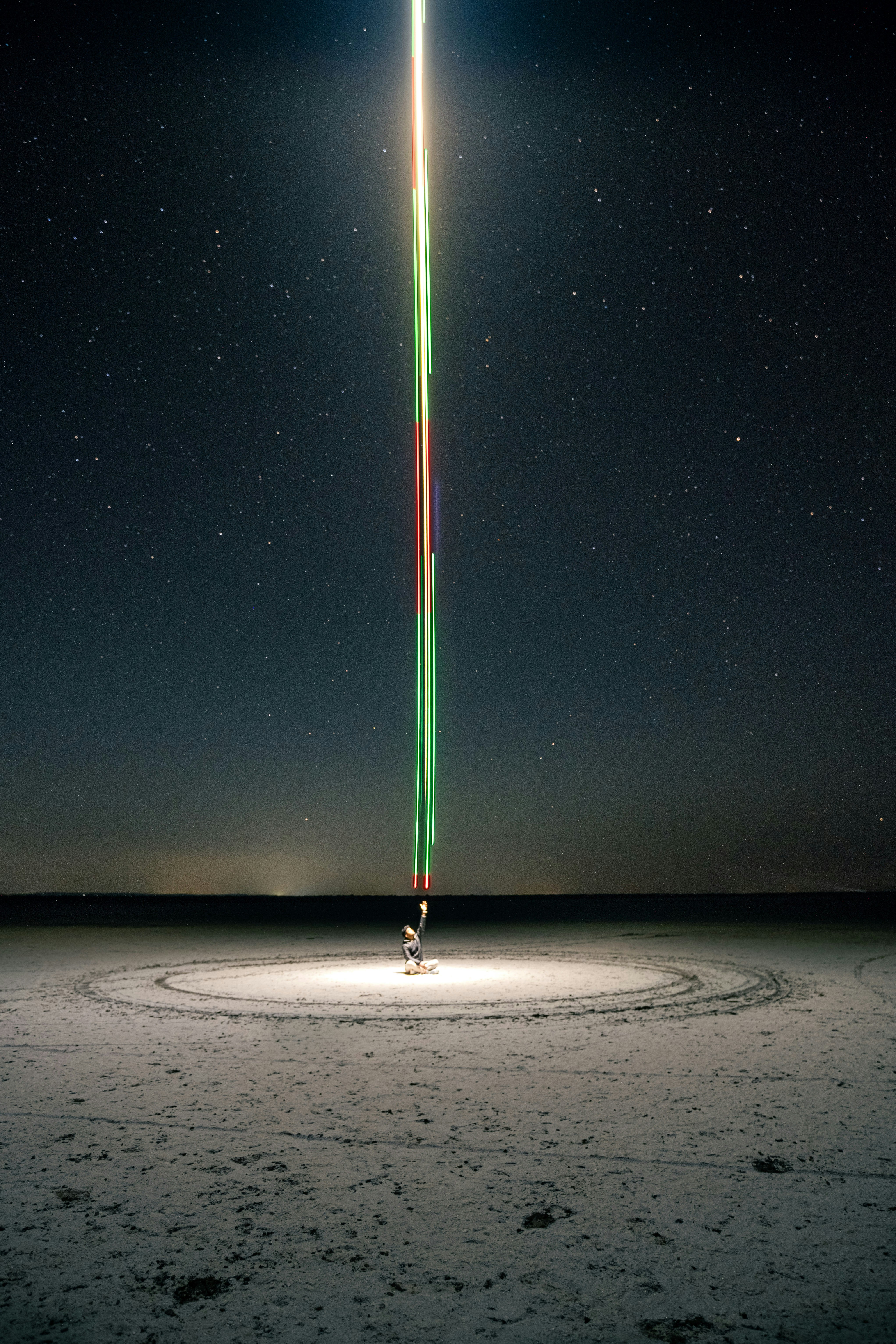 A solitary figure stands in a circular light pattern on a dark landscape, with vibrant colored beams shooting upwards into the starry night sky.
