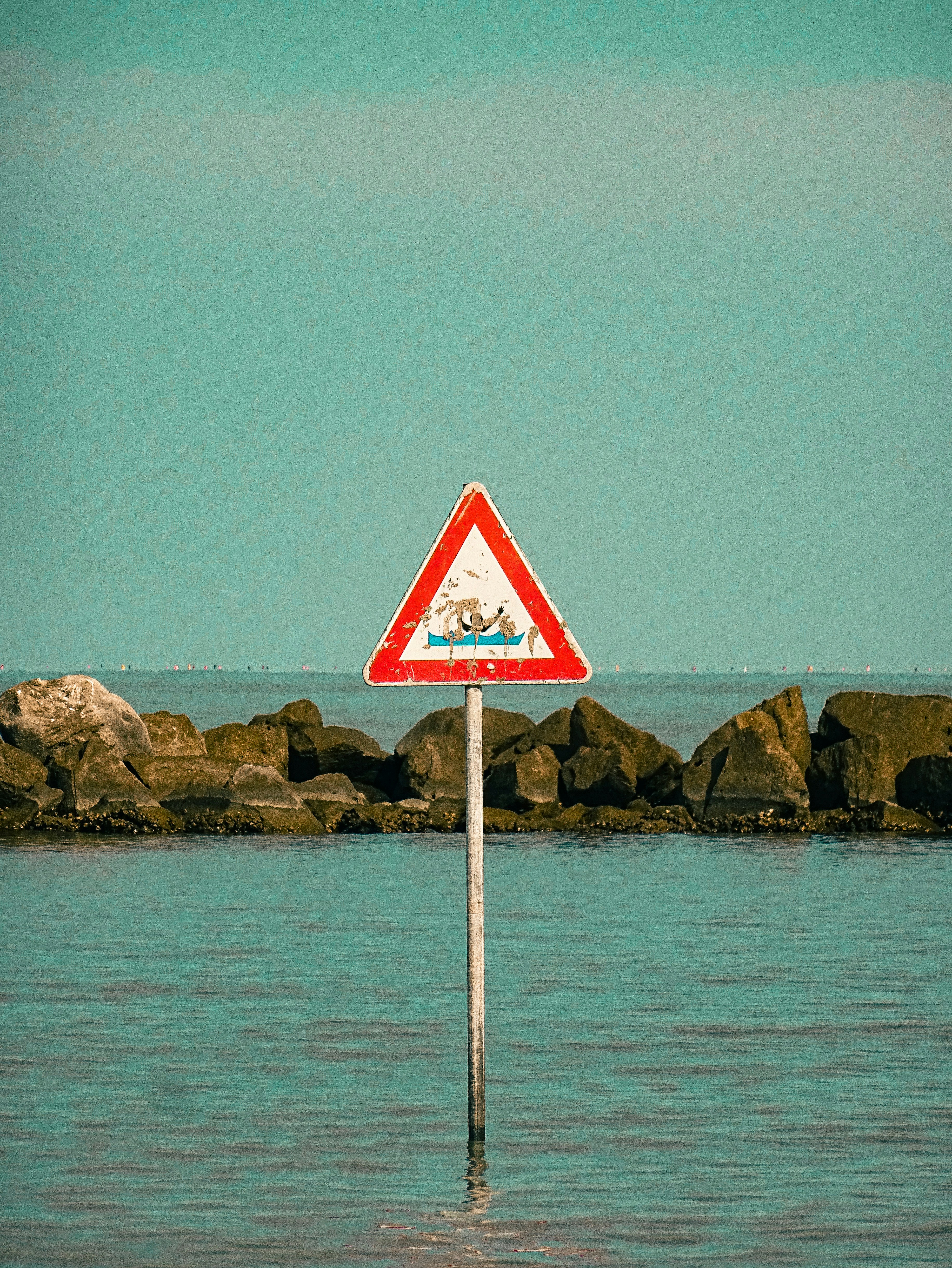 A weathered warning sign featuring a boat illustration stands tall in shallow water, surrounded by rocky formations. The serene backdrop complements the cautionary theme.