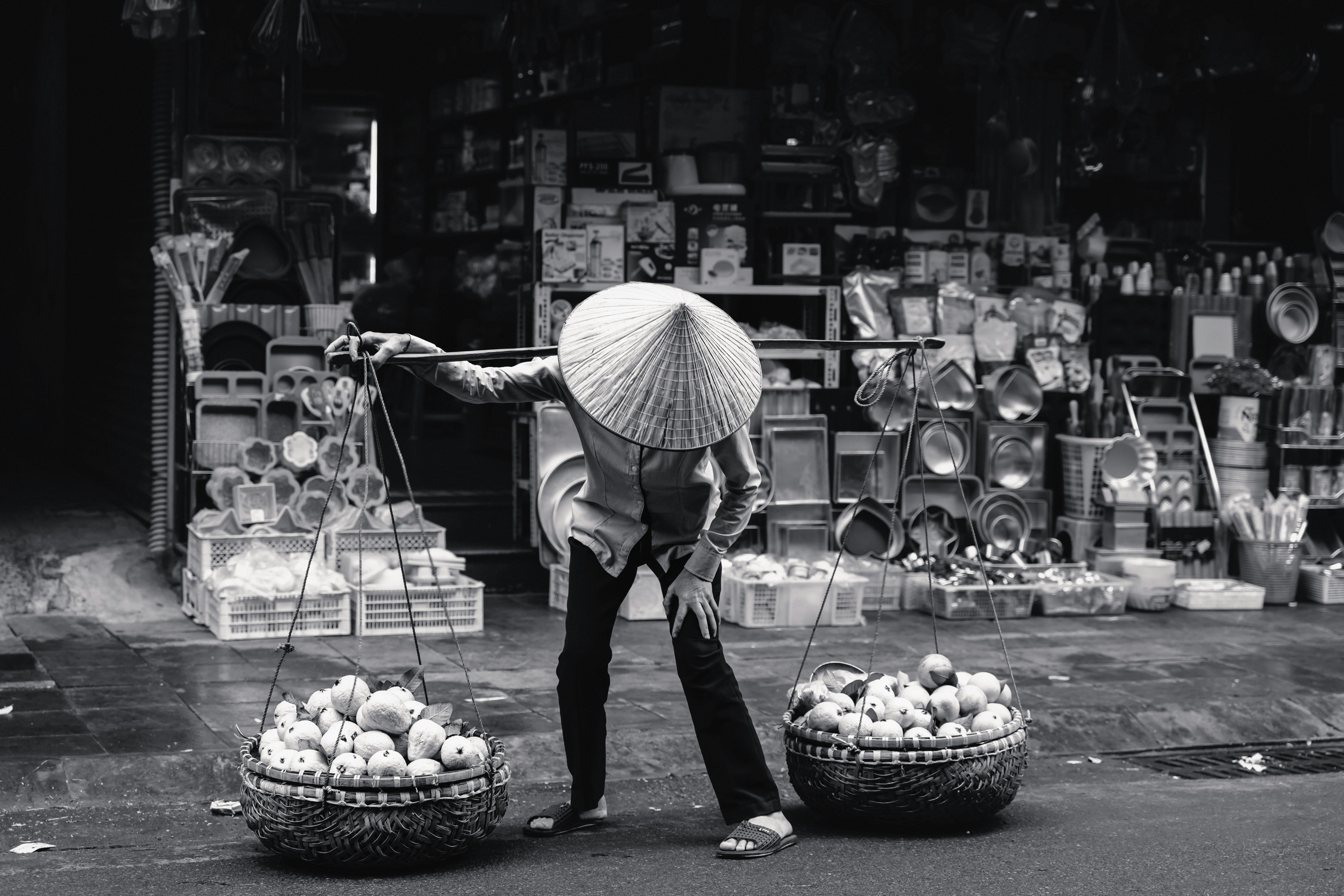 Street vendor with a conical hat balancing two baskets of fruit in front of a shop in Hanoi.