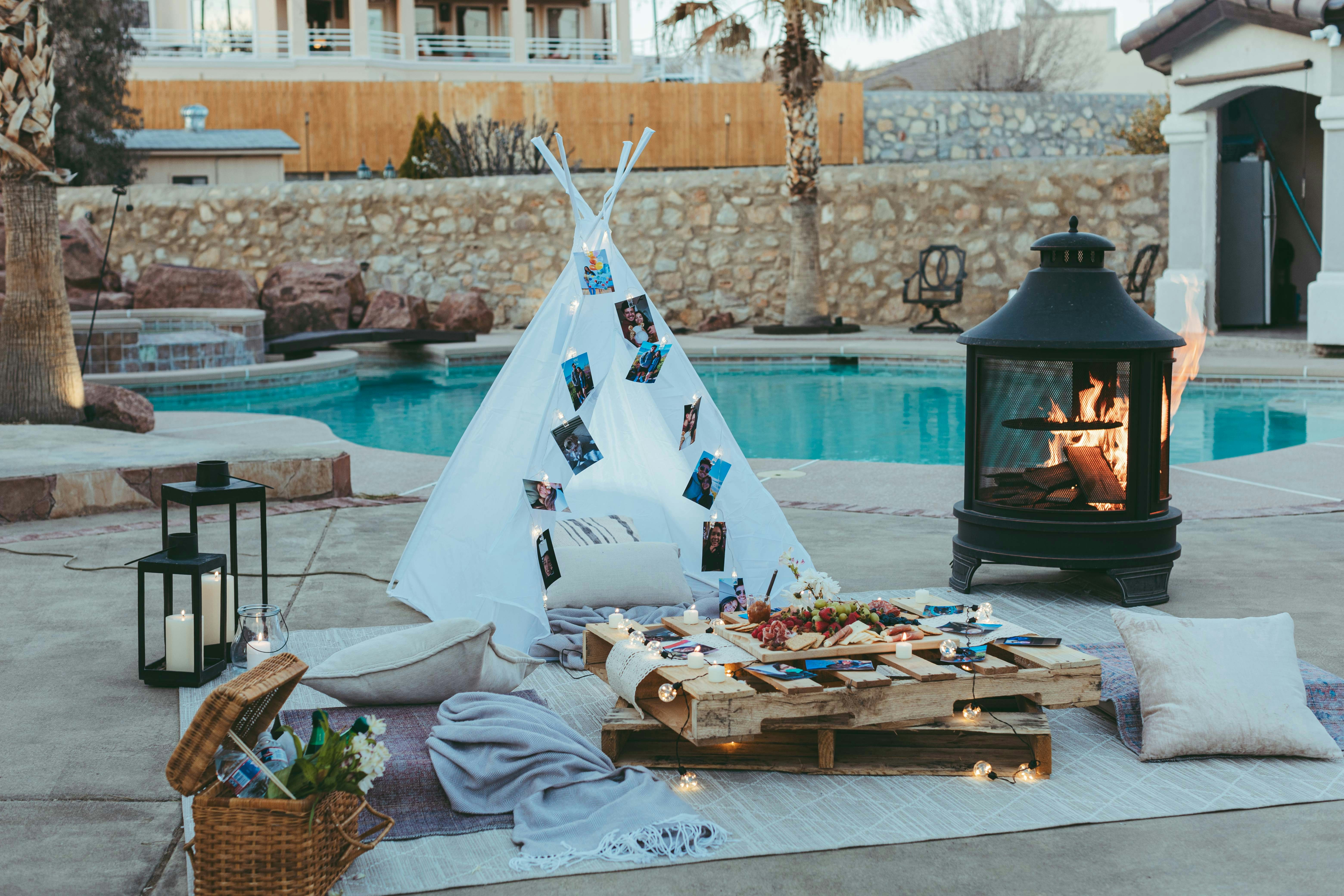 Outdoor setup featuring a teepee adorned with photos, a wooden table with a spread of snacks, and a warm fire pit, creating an inviting atmosphere for relaxation.