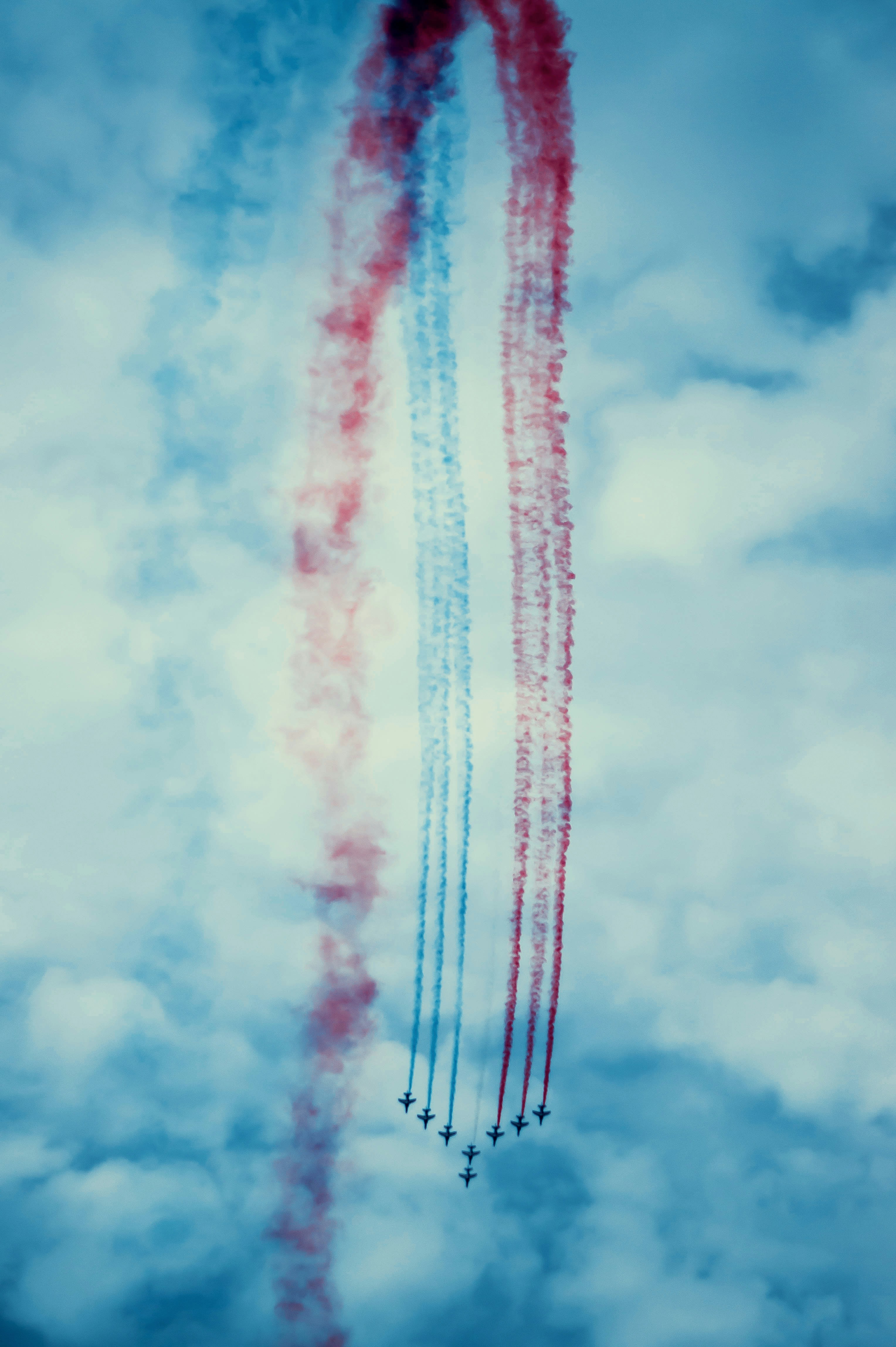Five jets fly in a tight formation, releasing red and blue smoke trails across a bright, cloud-filled sky. The photo highlights precise aerobatic choreography and vivid color contrast.