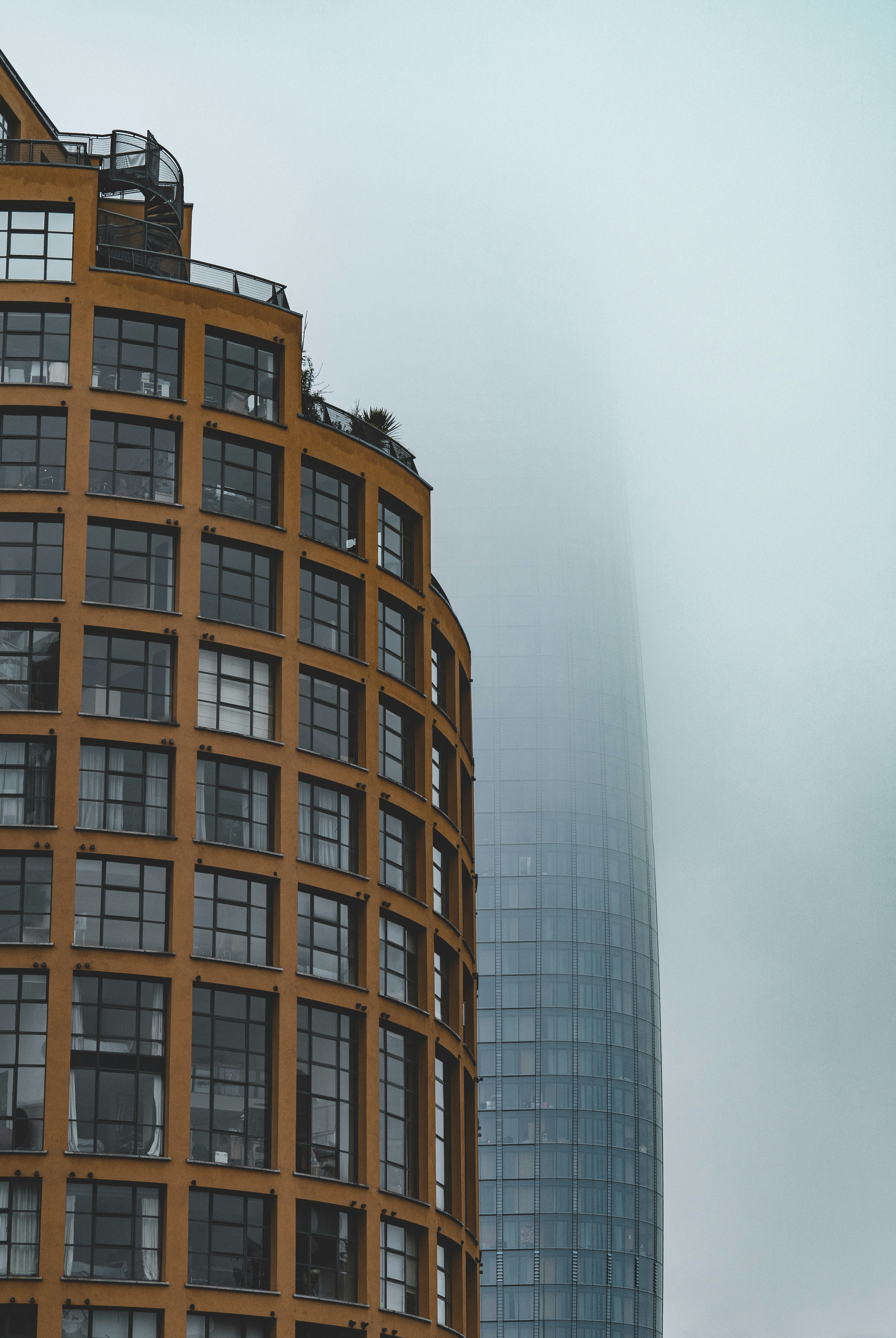 Rounded orange brick facade dominates the foreground as a glass skyscraper recedes into mist. The image juxtaposes brick texture with glass silhouette to convey a moody urban atmosphere.