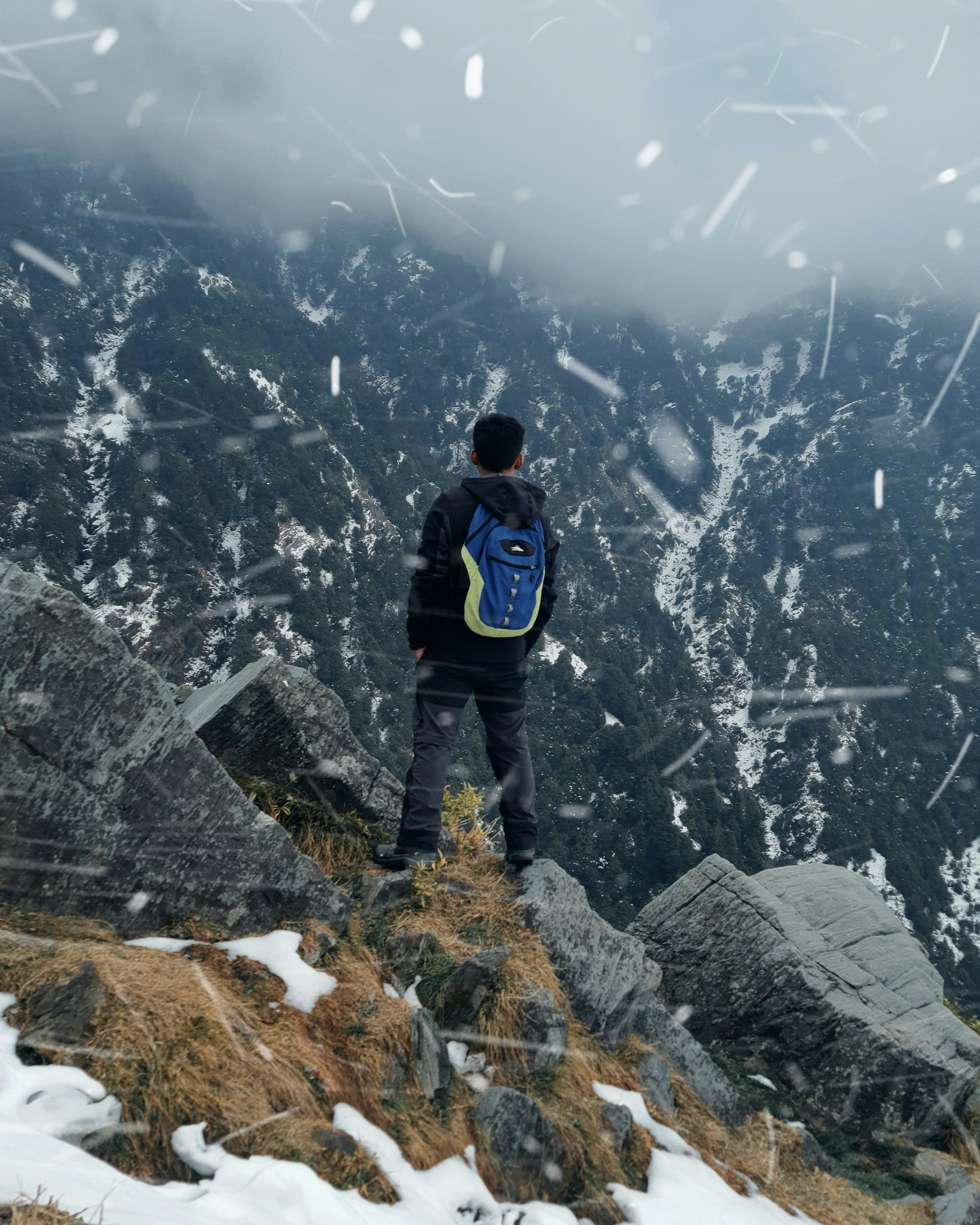 A lone hiker with a blue backpack stands on a jagged, snow-dusted ridge overlooking a forested valley as snow falls.