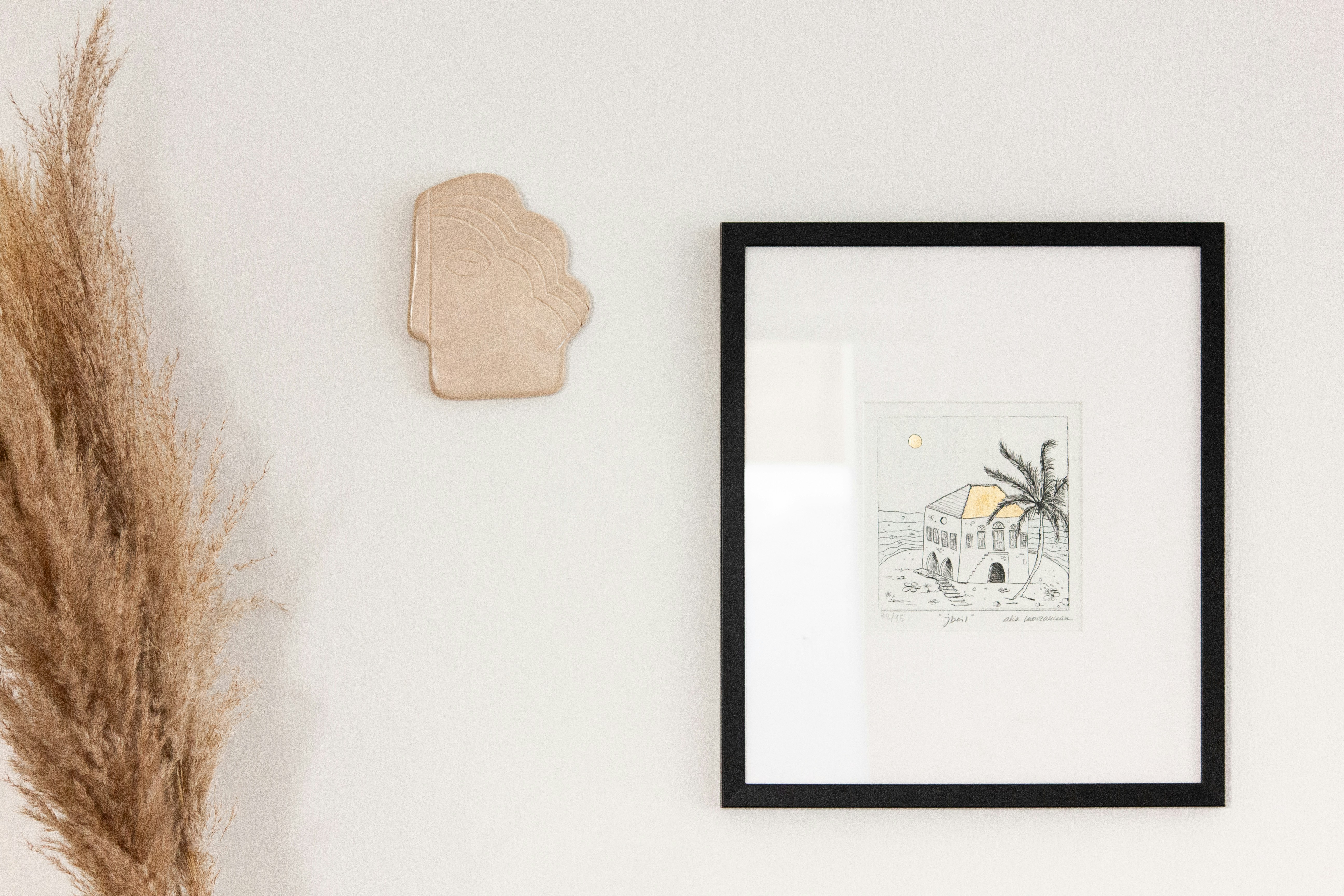 Minimalist wall decor featuring a framed illustration of a coastal scene alongside a sculptural wall piece, complemented by soft pampas grass.