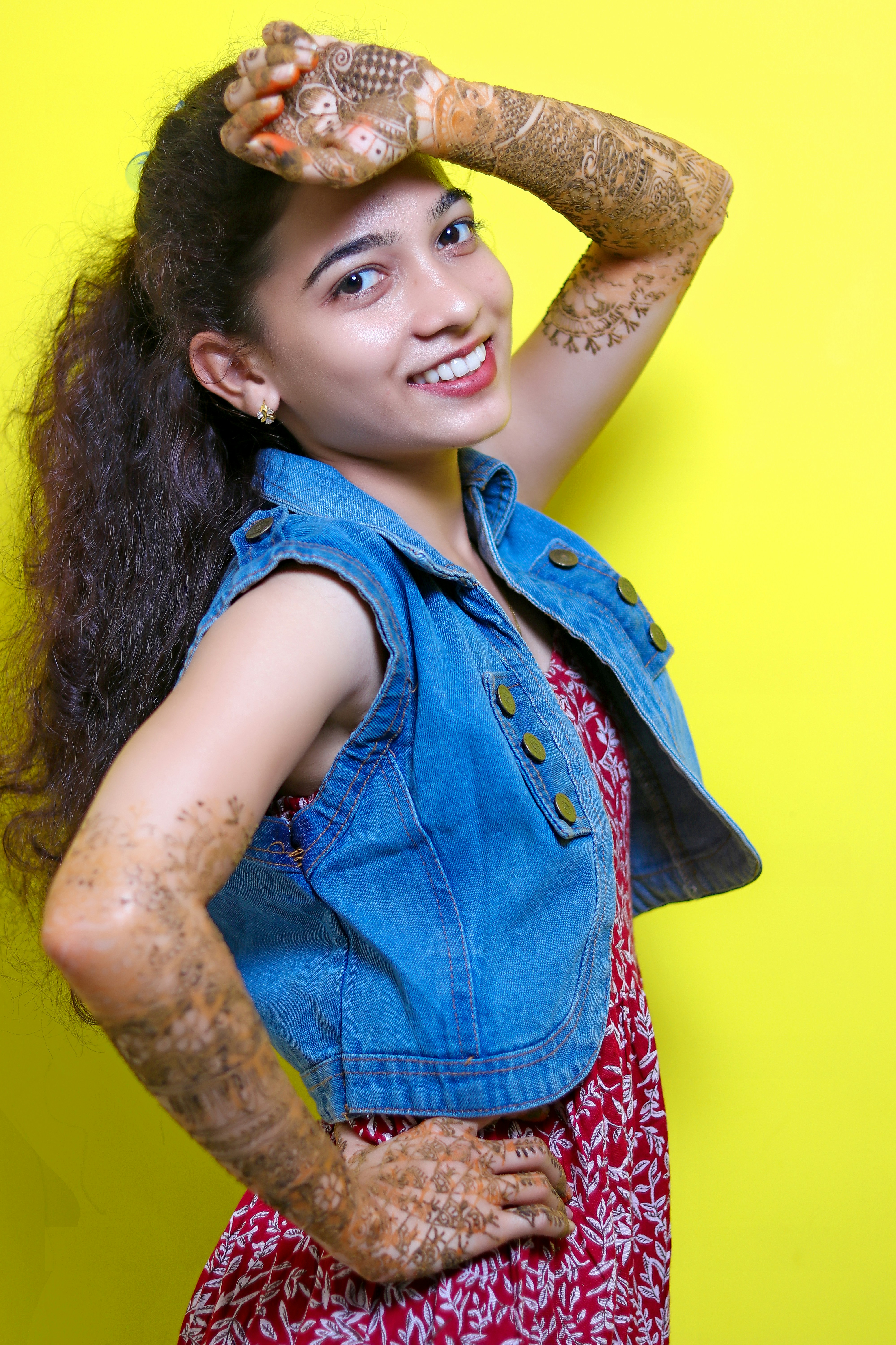 A young woman poses confidently against a bright yellow background, showcasing intricate henna designs on her arms while wearing a denim vest over a floral dress.