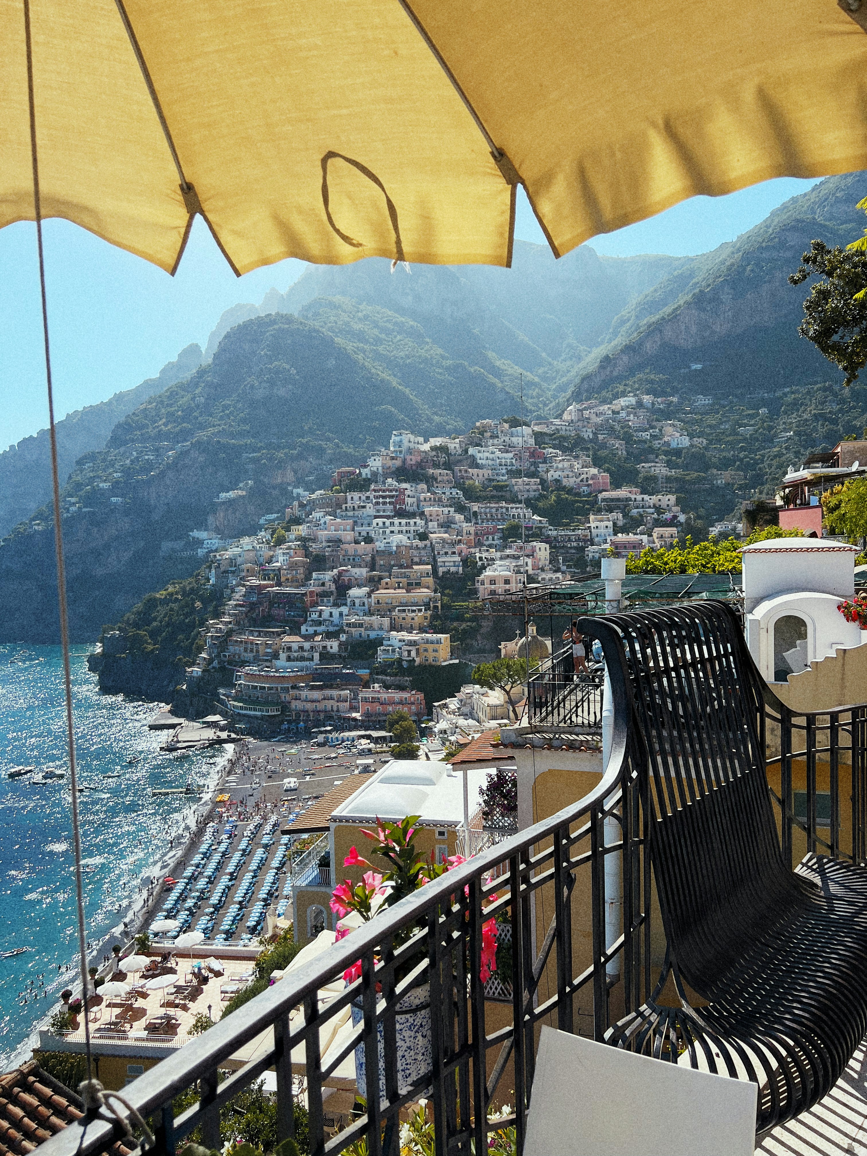 Vibrant coastal village nestled among lush mountains, showcasing colorful buildings and a serene beach below. A sunshade and balcony frame the view.