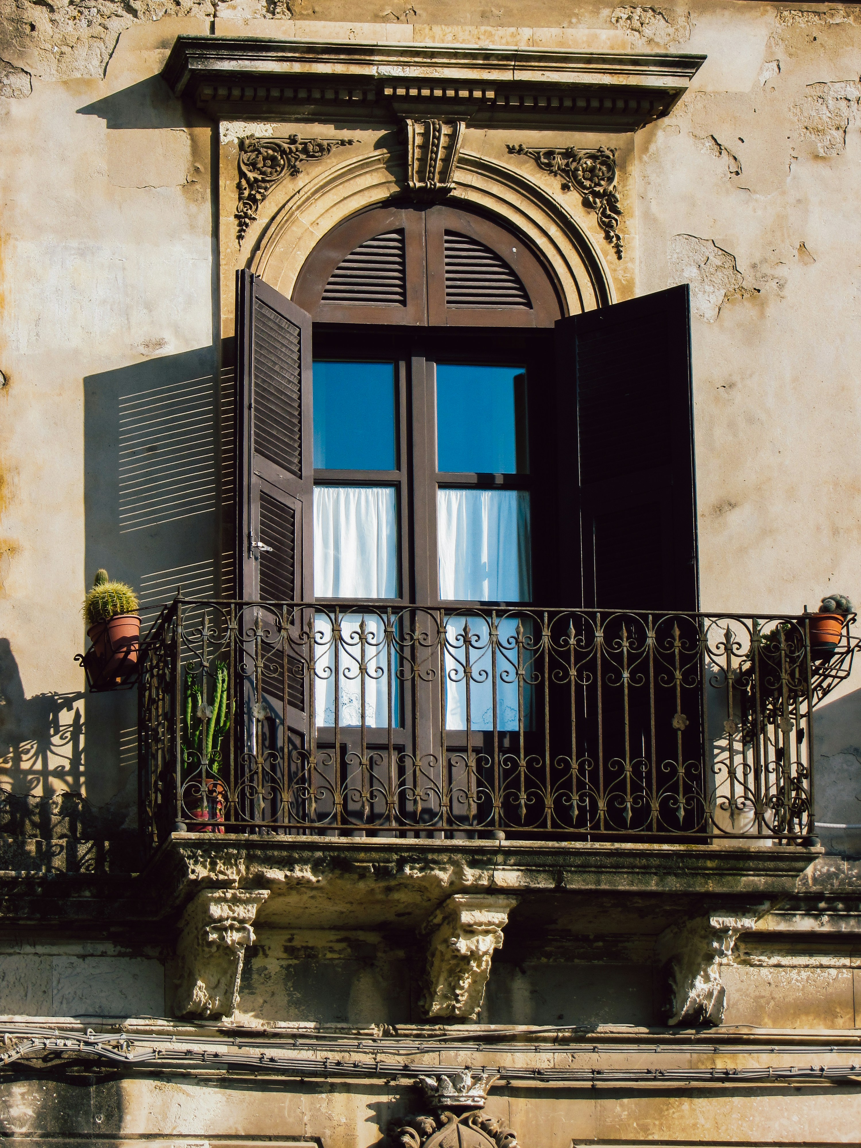 A weathered balcony with open shutters reveals a glimpse of white curtains, framed by ornate architecture and potted plants. 