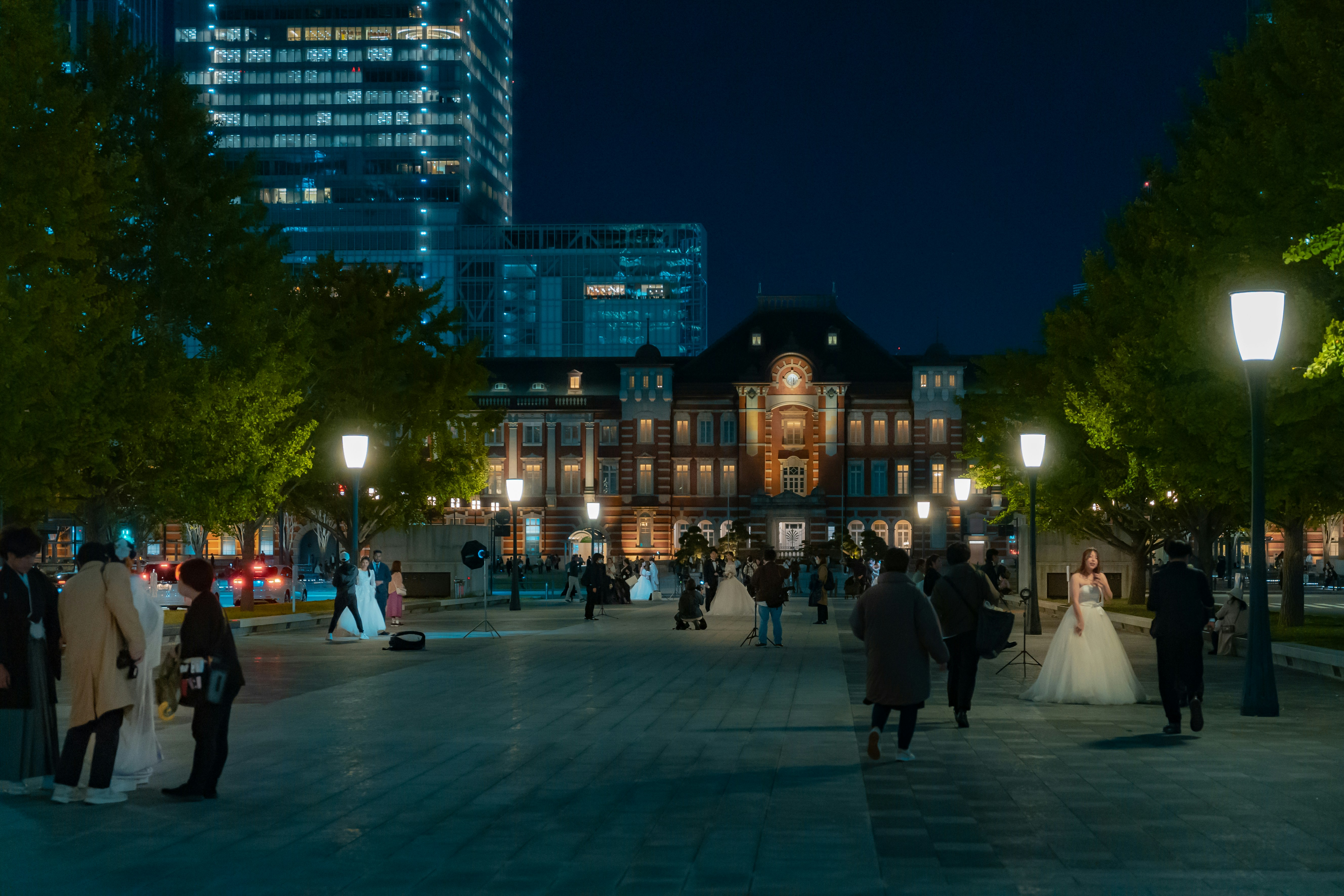 Couples in formal attire stroll through a beautifully lit plaza near a historic building at night. The scene captures a blend of modernity and tradition.