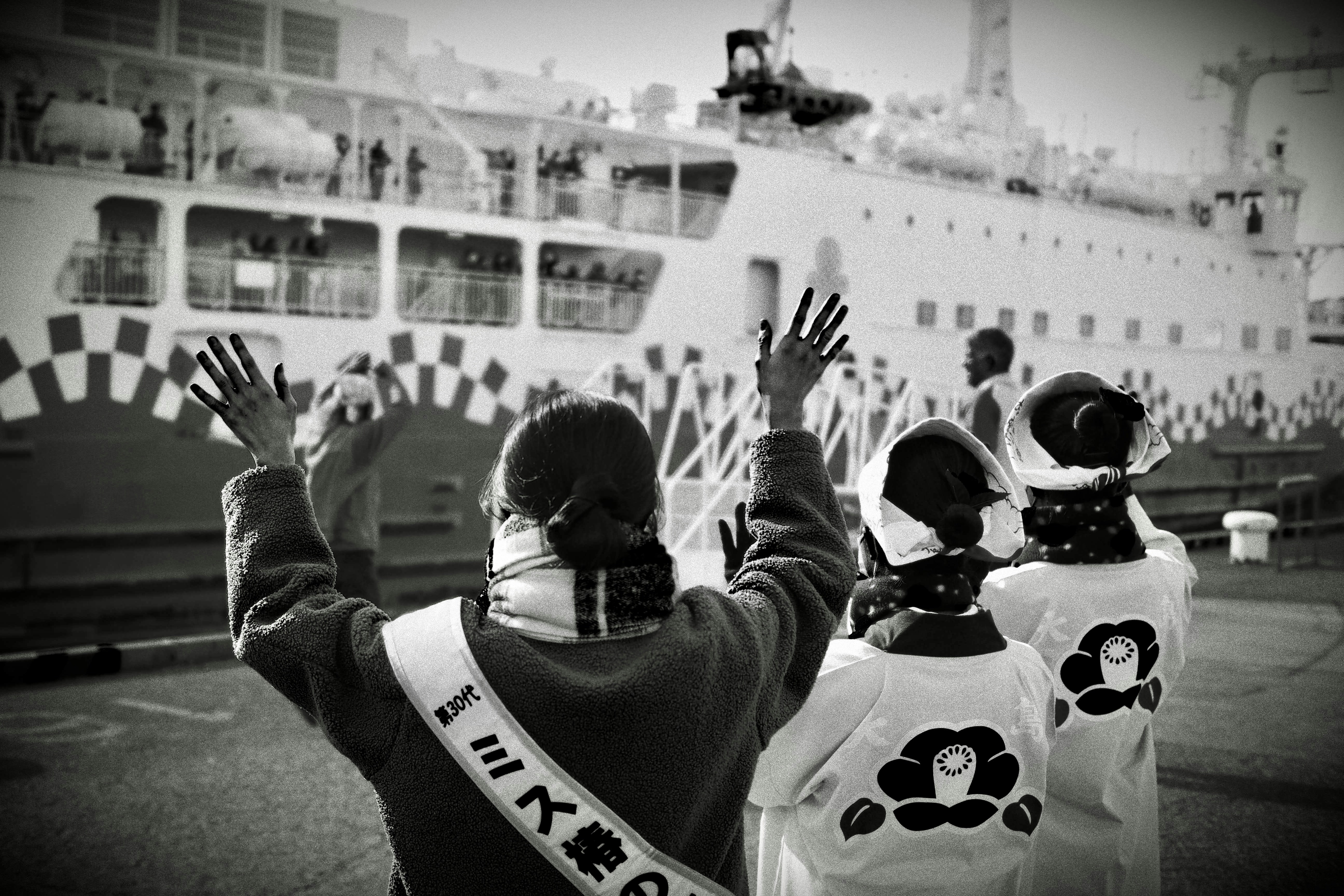 Three individuals in traditional attire wave goodbye to a departing ship, capturing a moment of connection and emotion. The scene is rendered in black and white, emphasizing the poignant atmosphere.