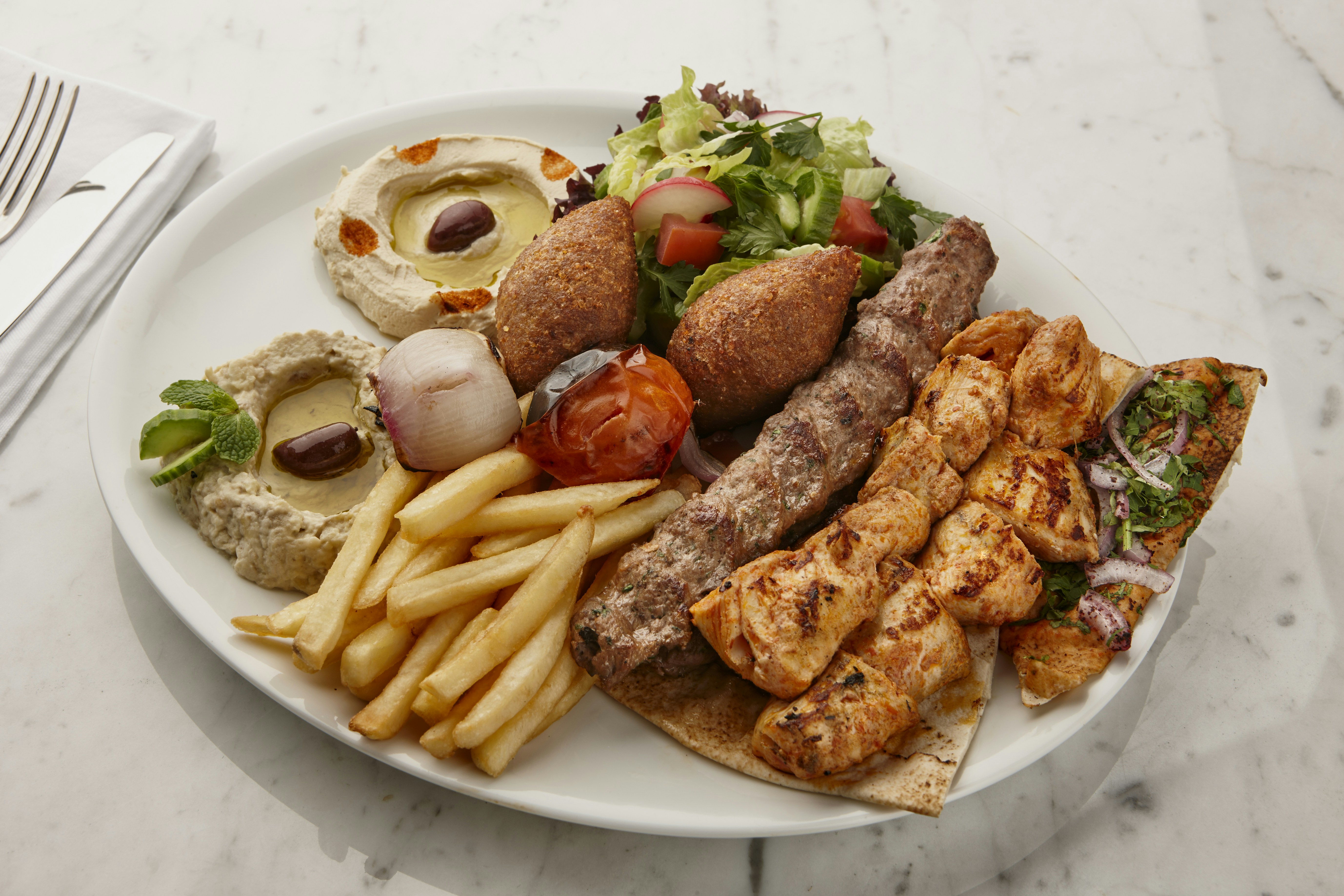 Plate of grilled kebabs, pita, hummus with olive, fries, and a fresh salad on a white marble table.