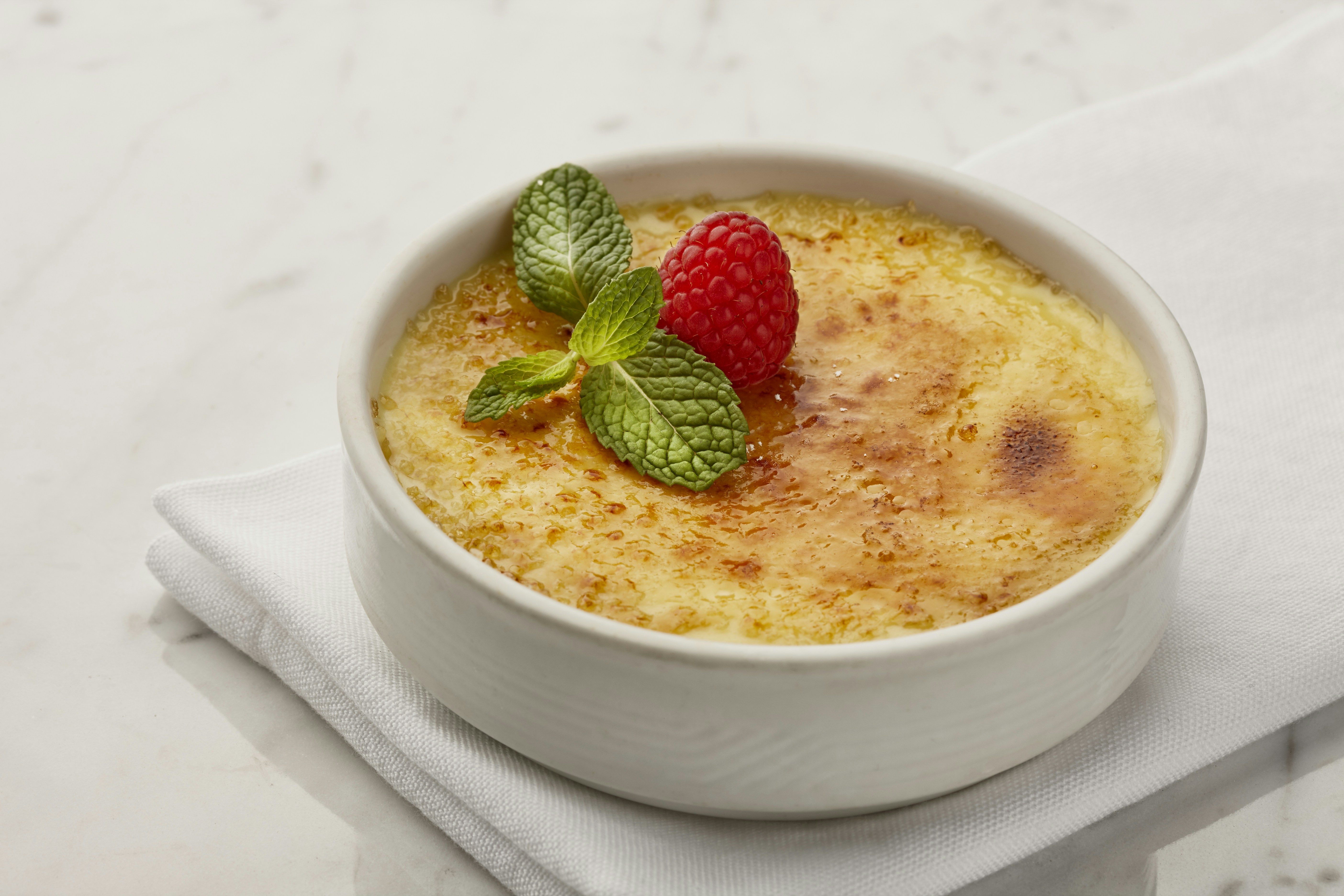 Crème brûlée topped with fresh raspberries and mint leaves, elegantly presented on a white cloth.