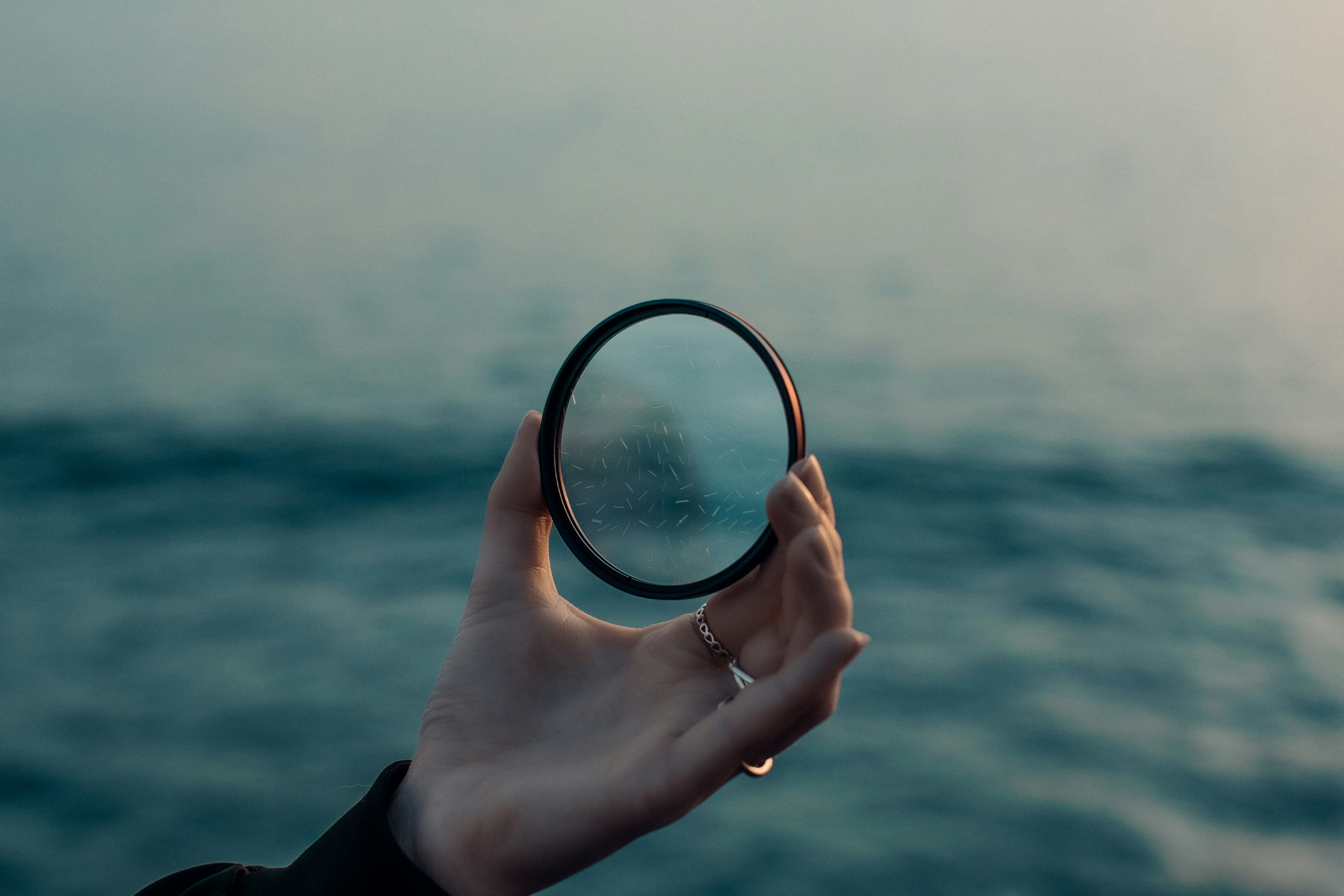 Hand holding a camera filter against a blurred ocean background.