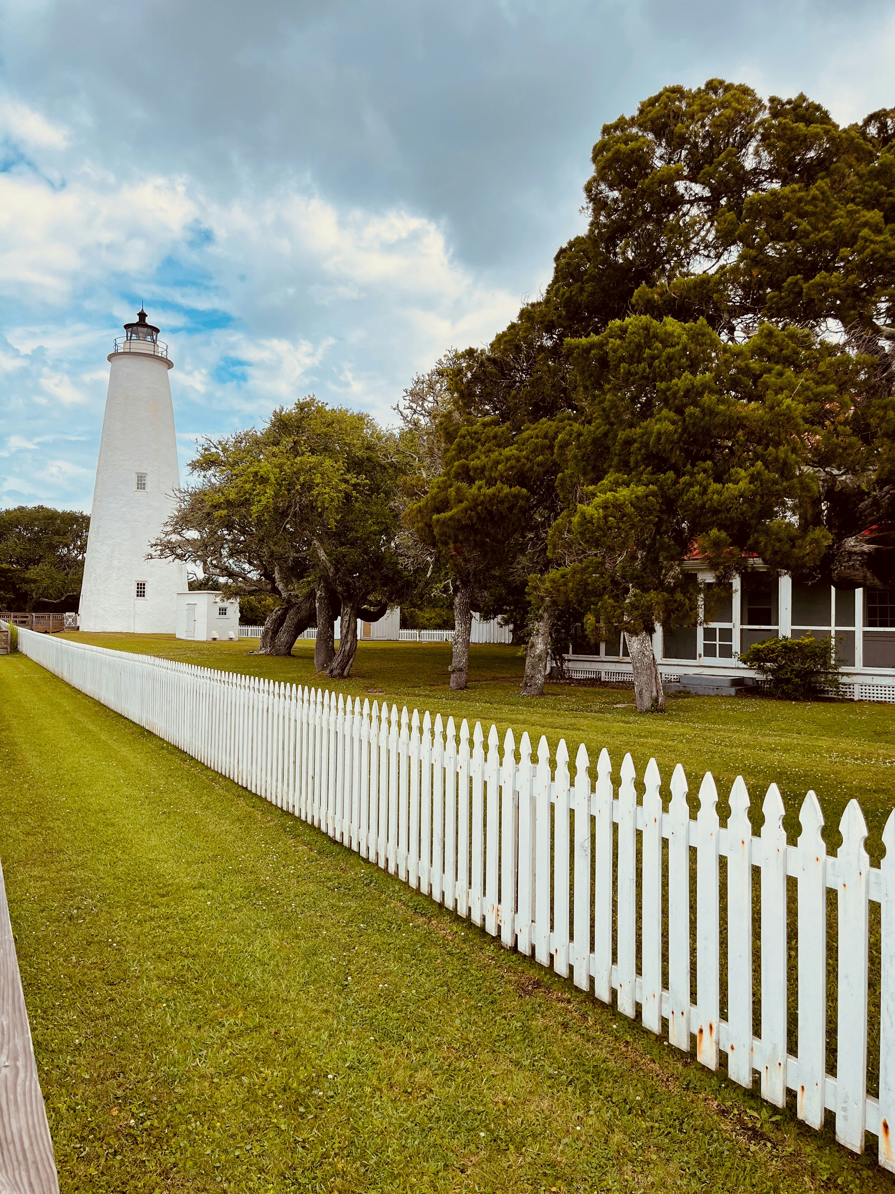 A historic lighthouse stands tall amidst lush greenery and a white picket fence, showcasing a serene coastal landscape.