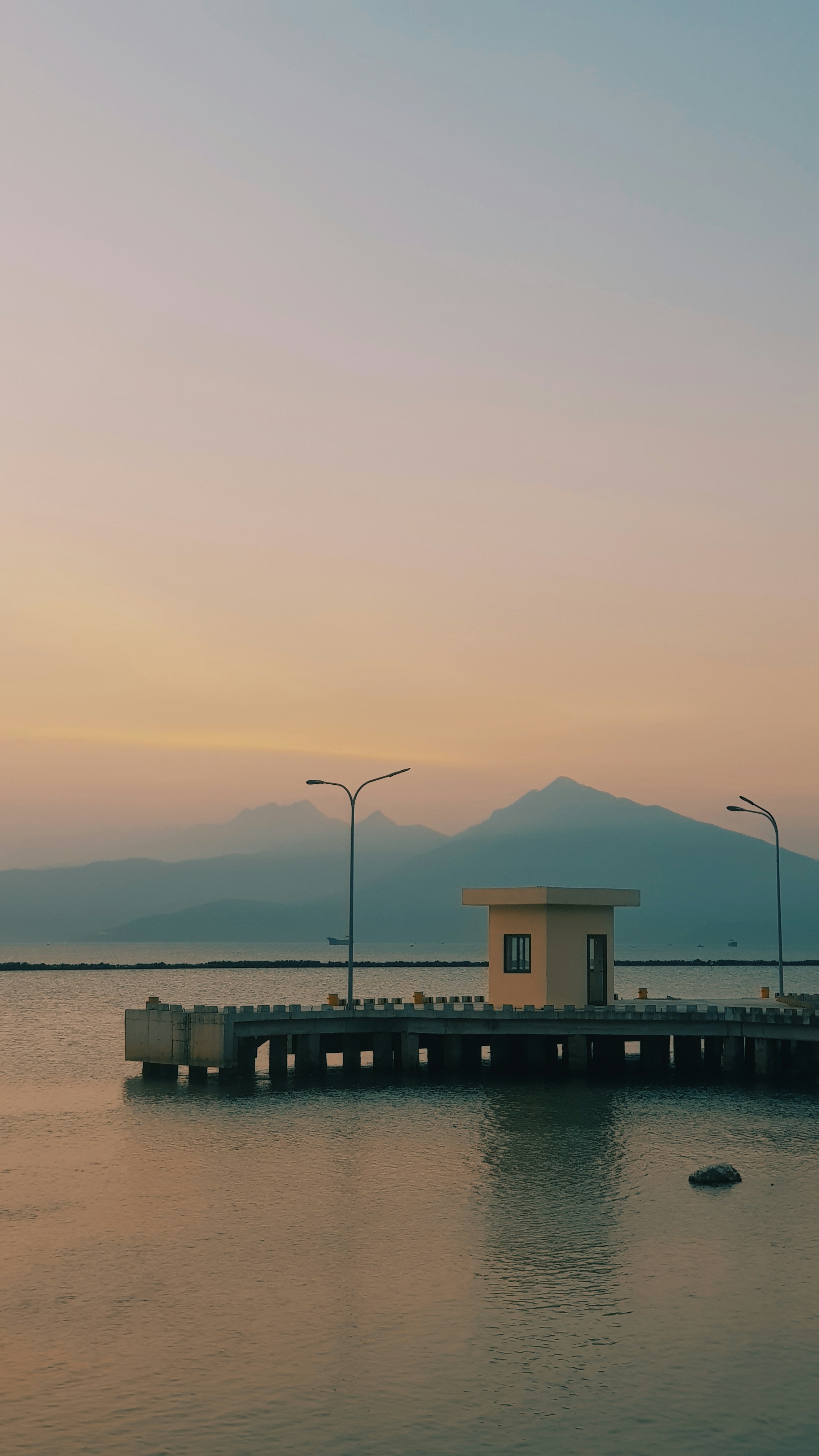 Small coastal structure on a pier with gentle waves and mountains in the background during sunset.