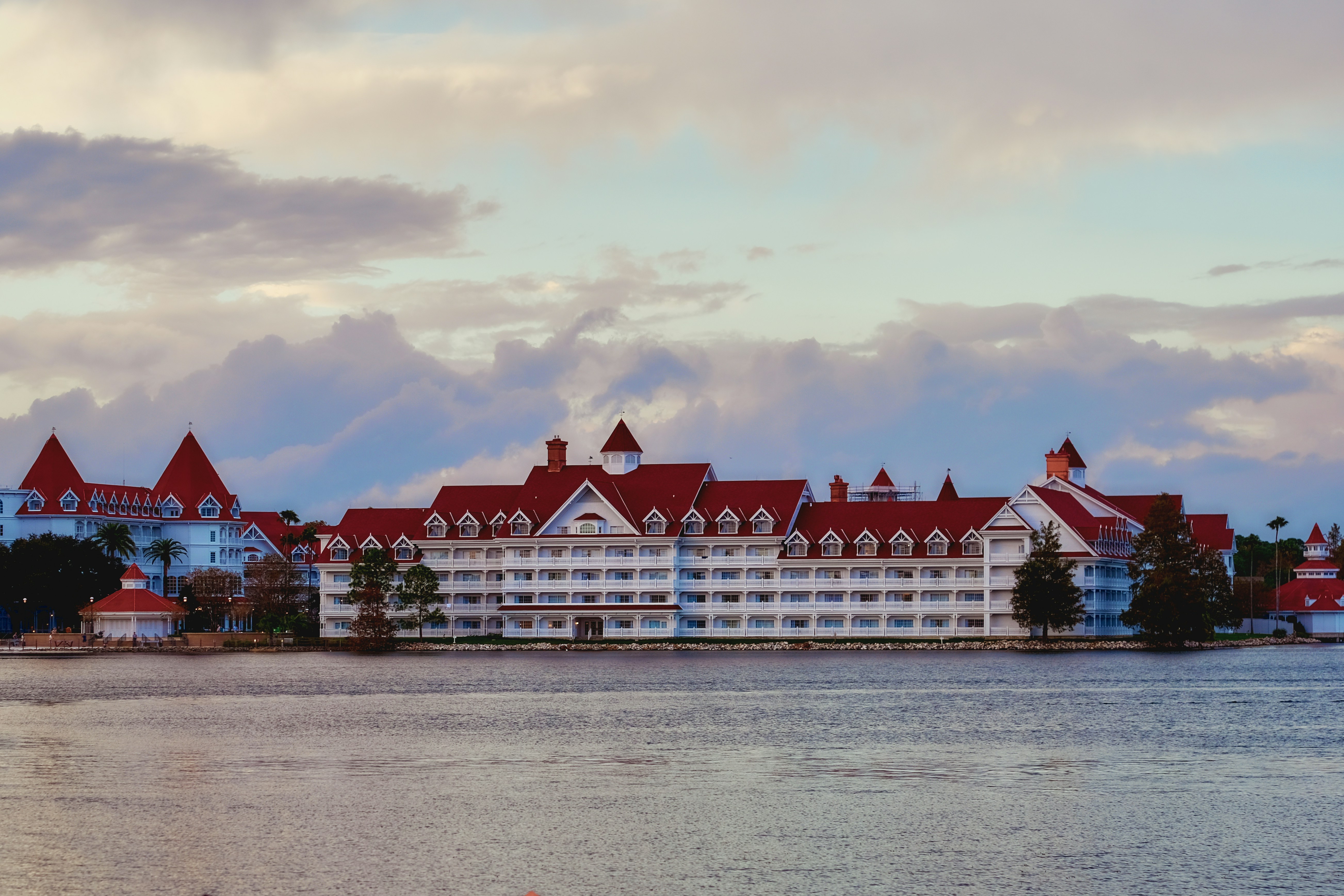Large building with red roofs adjacent to a calm body of water under a cloud-streaked sky.