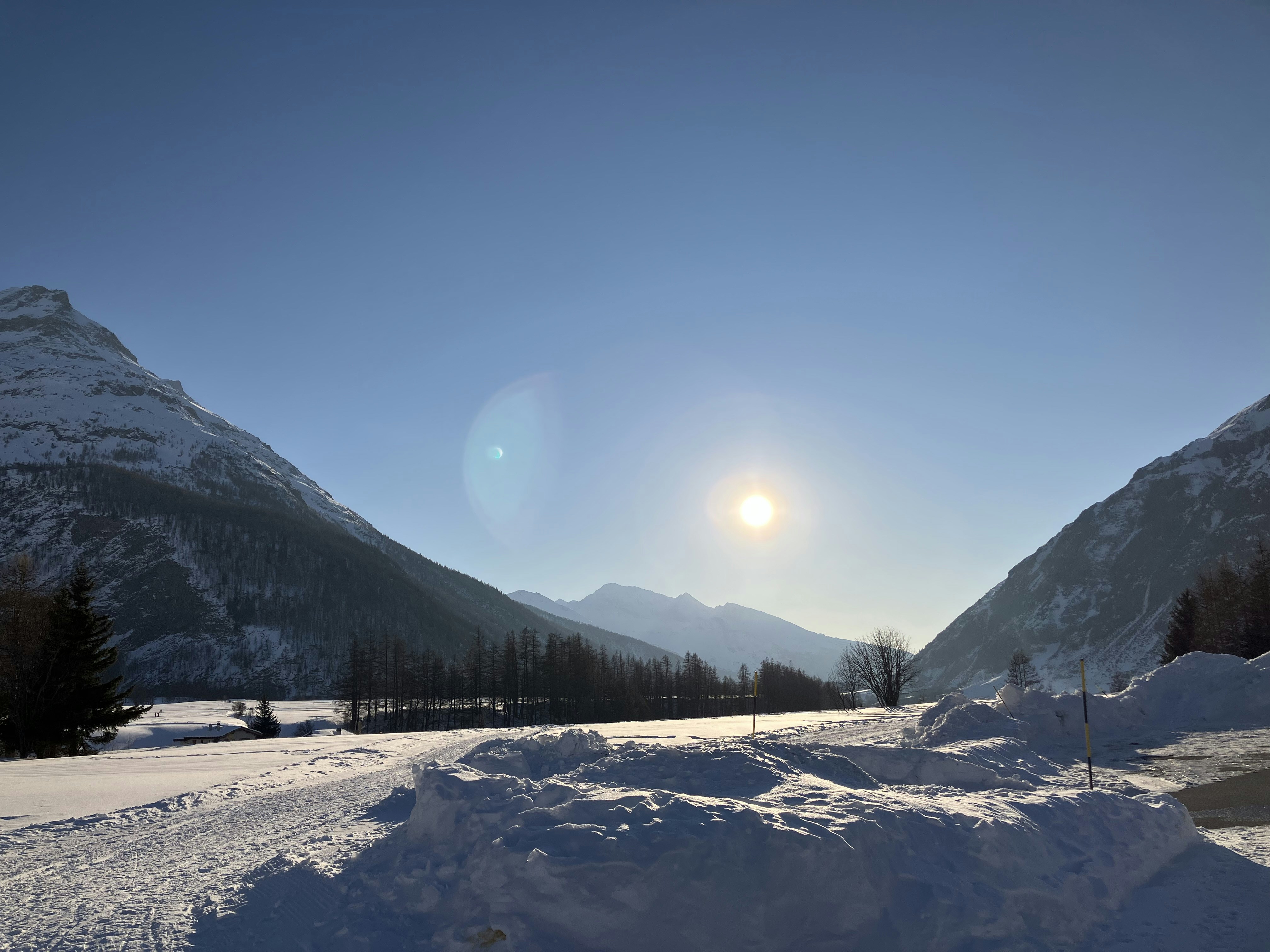 Snow-covered valley flanked by majestic mountains under a clear blue sky with a bright sun illuminating the scene.