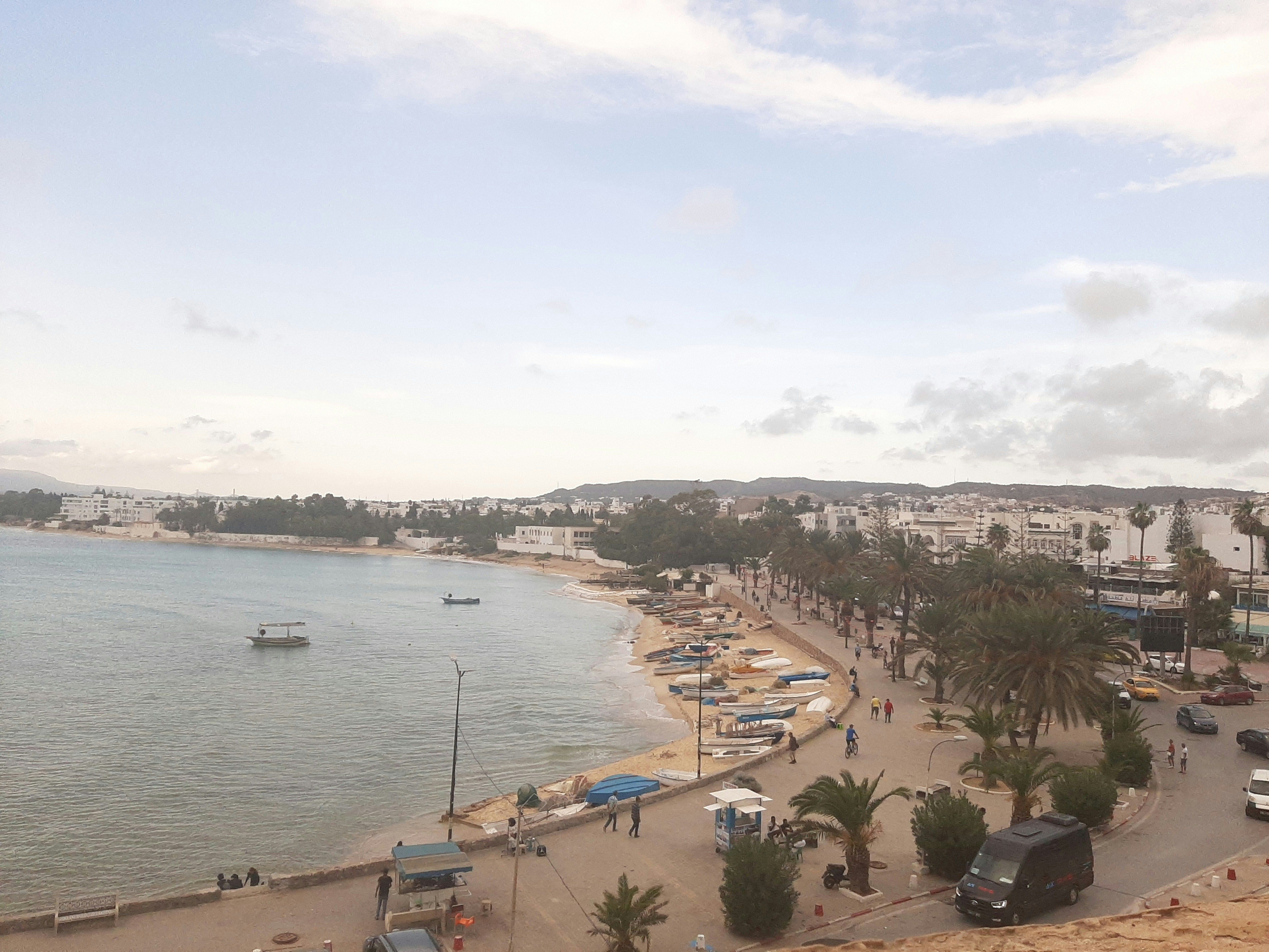 Coastal scene featuring a sandy beach lined with palm trees and boats, with people strolling along the promenade. The backdrop includes white buildings under a cloudy sky.