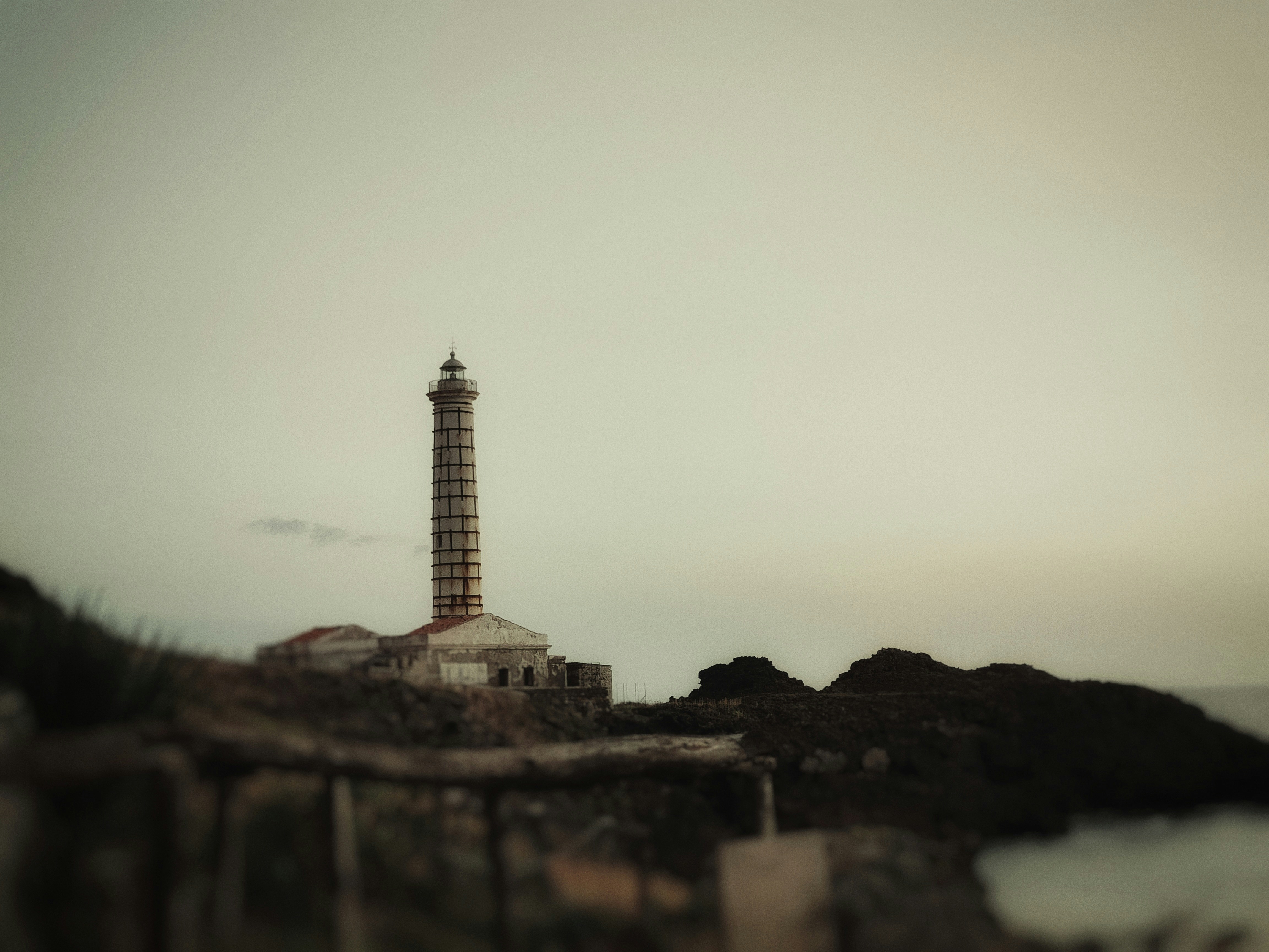 Muted vintage-toned photograph of a solitary lighthouse on a jagged shoreline, under a pale, expansive sky. The composition emphasizes the lighthouse as the quiet focal point amid rugged rocks.