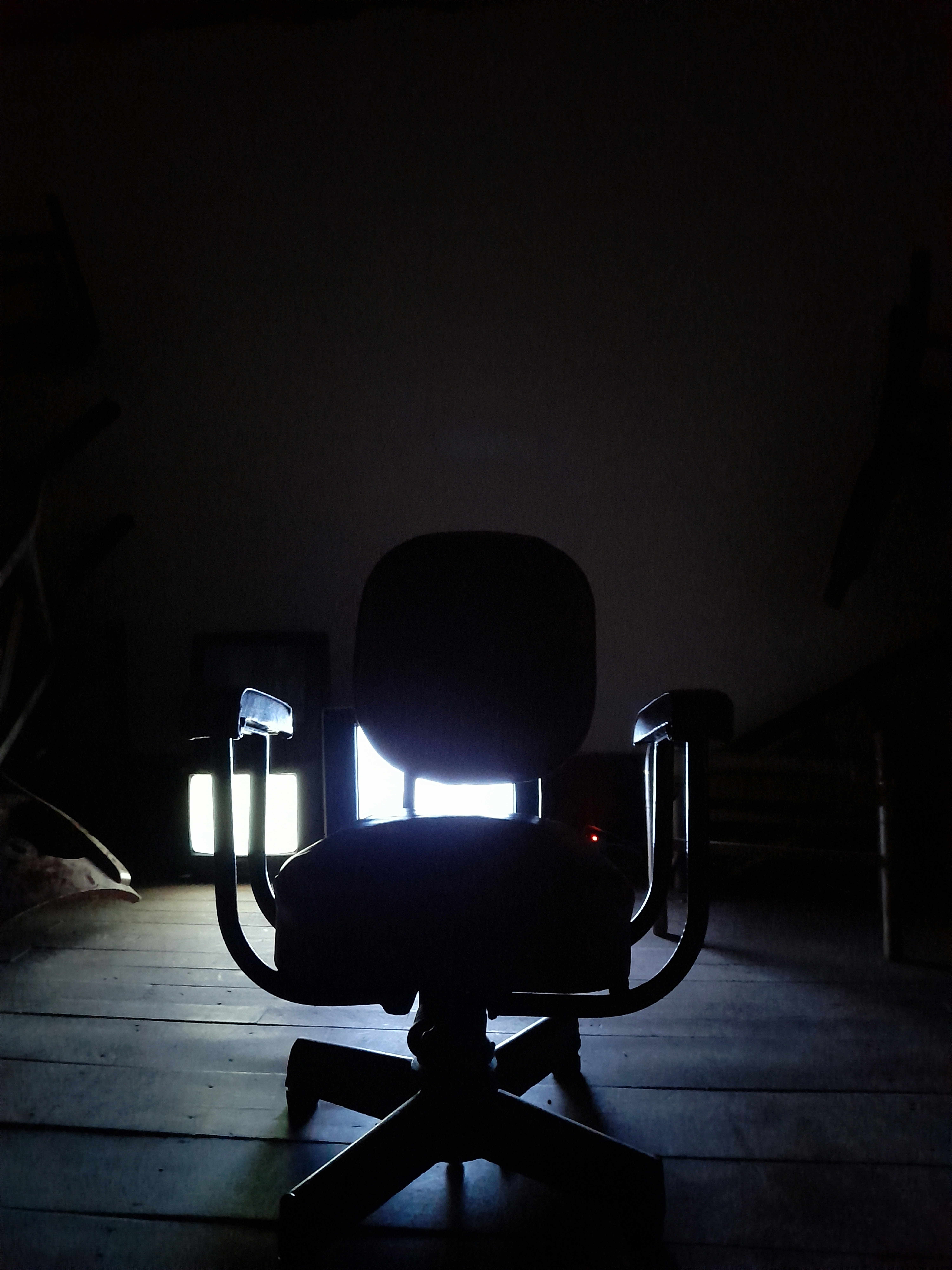 A solitary office chair sits in a dimly lit room, illuminated by the glow of two monitors behind it. The contrast between light and shadow creates a moody atmosphere.