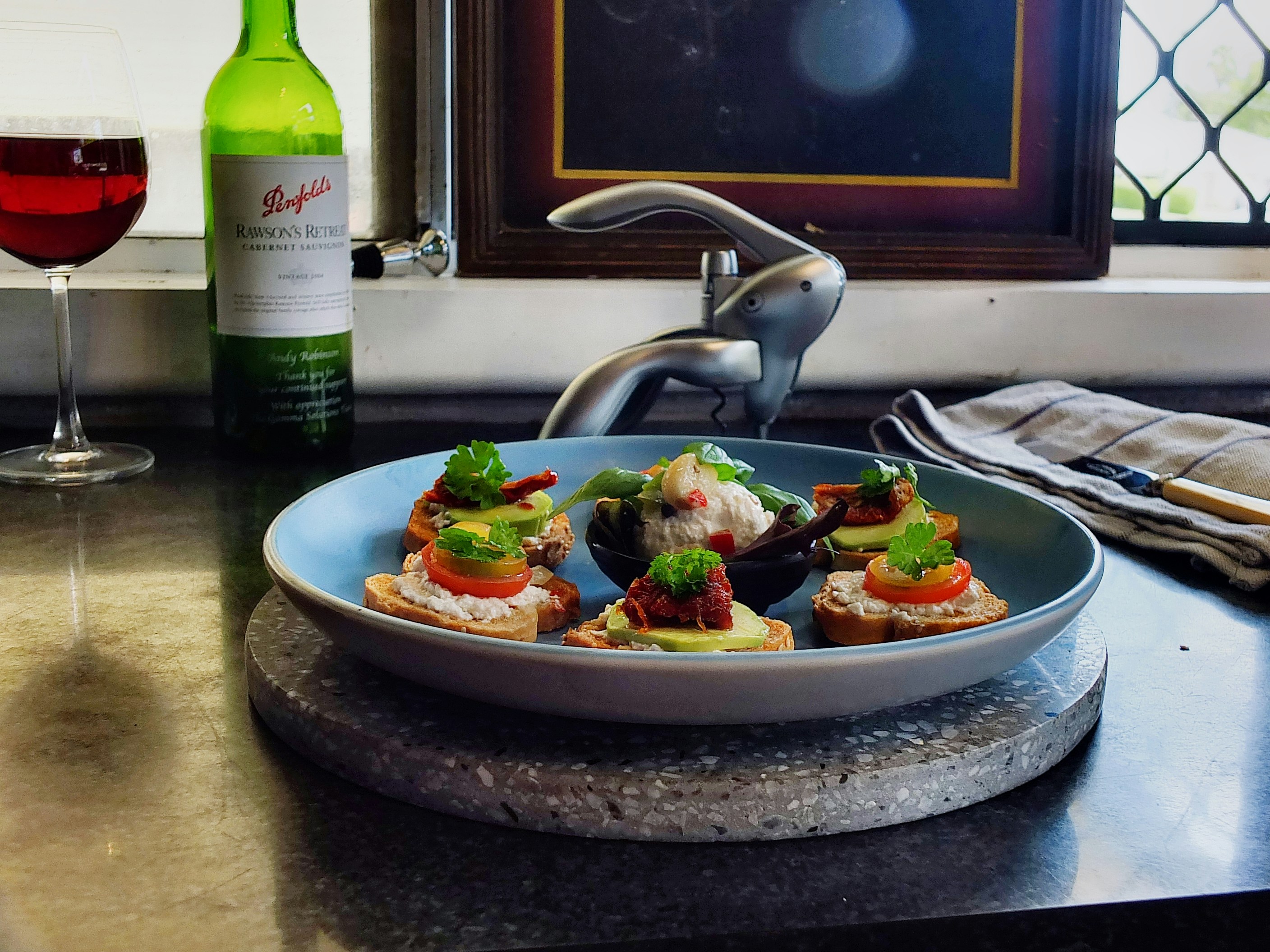 A beautifully arranged platter of gourmet canapés featuring vibrant toppings, accompanied by a glass of red wine and a bottle in the background. The setting showcases a modern kitchen ambiance.