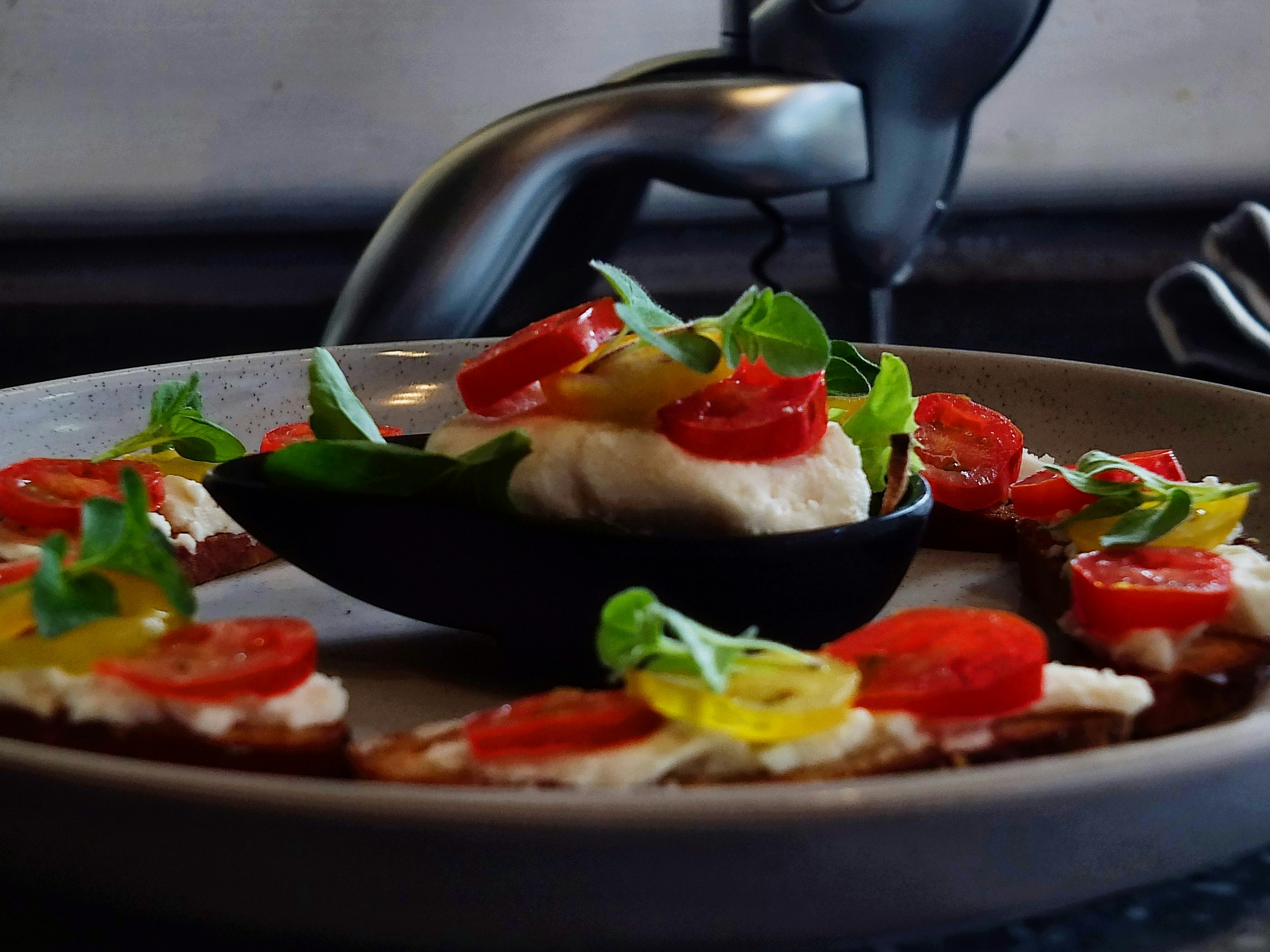 A skillet-style plate features a central spoon with herb-garnished dollop of cream surrounded by tomato-topped crostini, bright greens adding contrast.